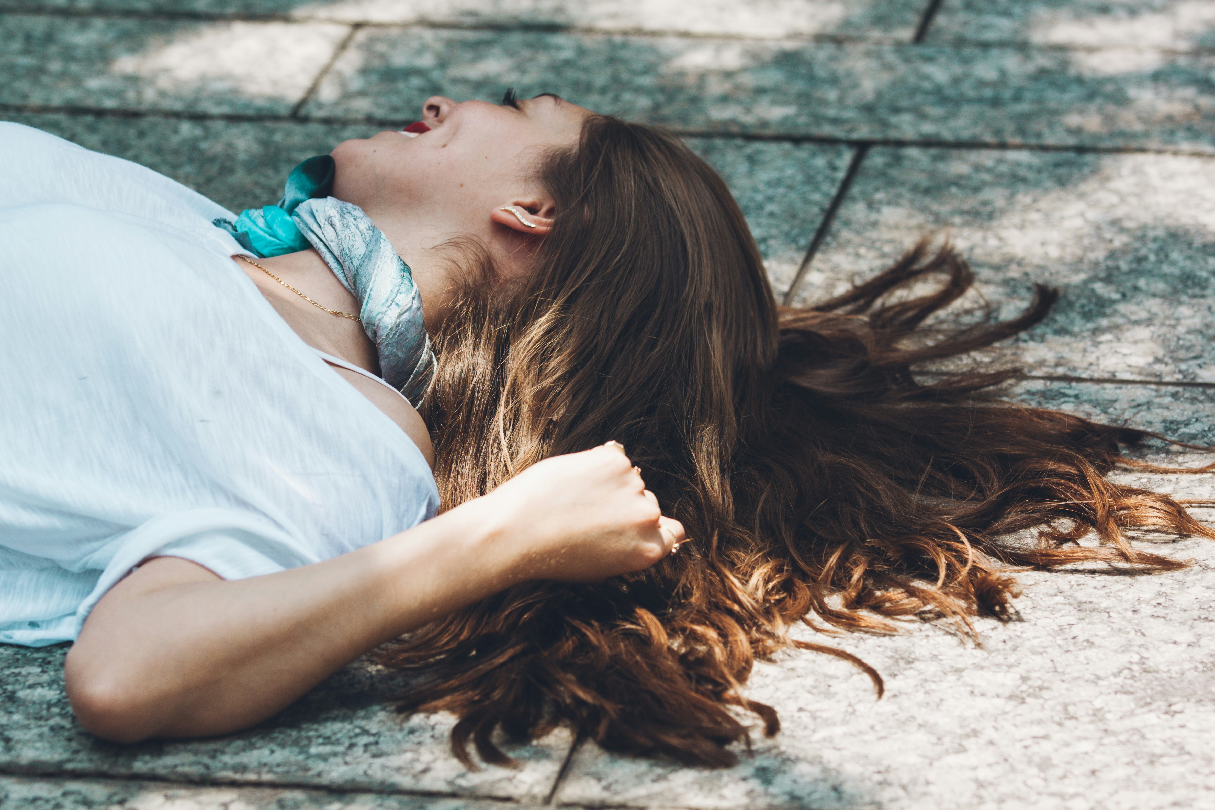 Woman lying on stone pavement with long hair splayed out, bathed in dappled sunlight.