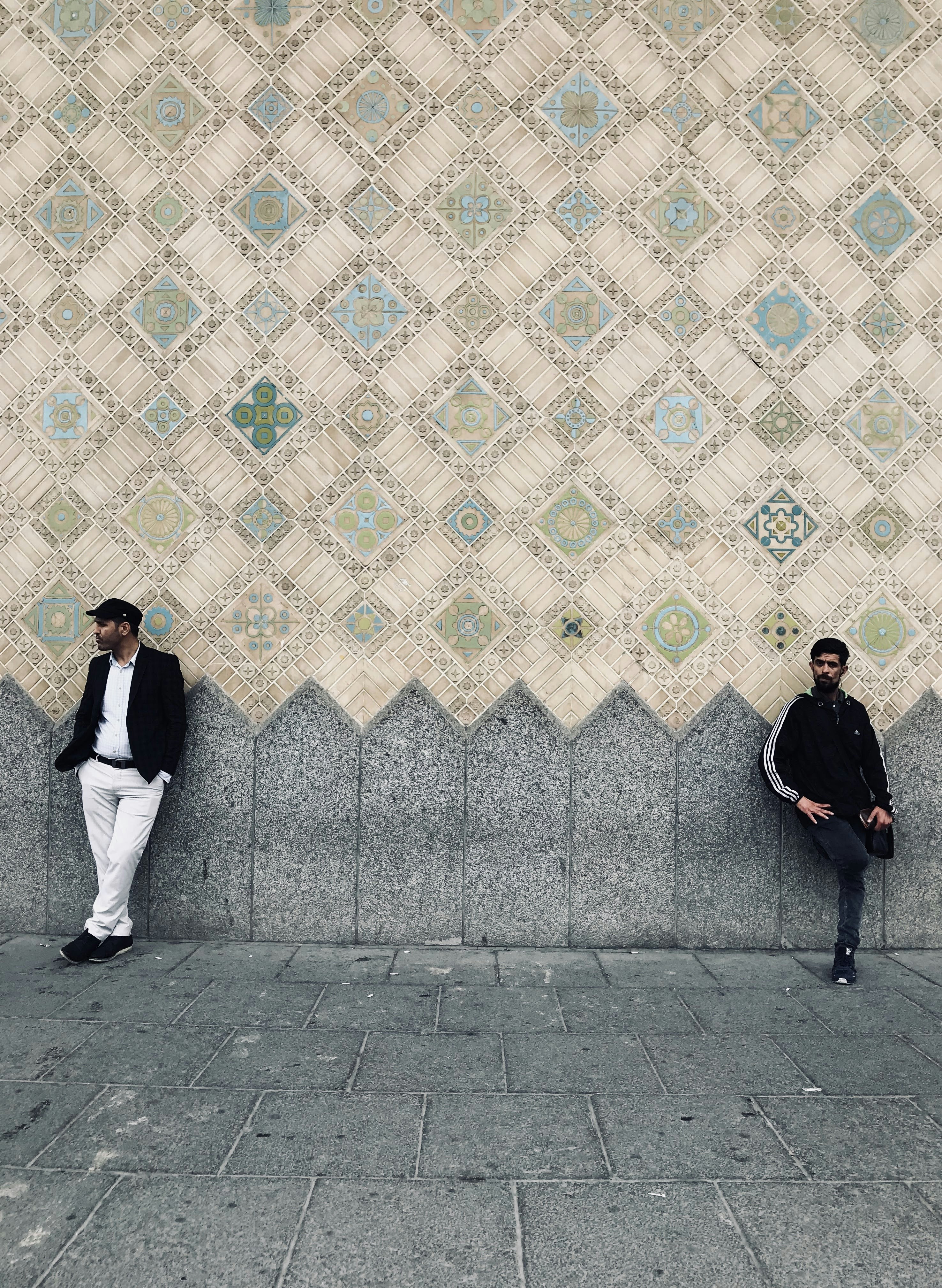 Two men leaning against a textured wall adorned with geometric patterns, showcasing a blend of modern style and traditional design.
