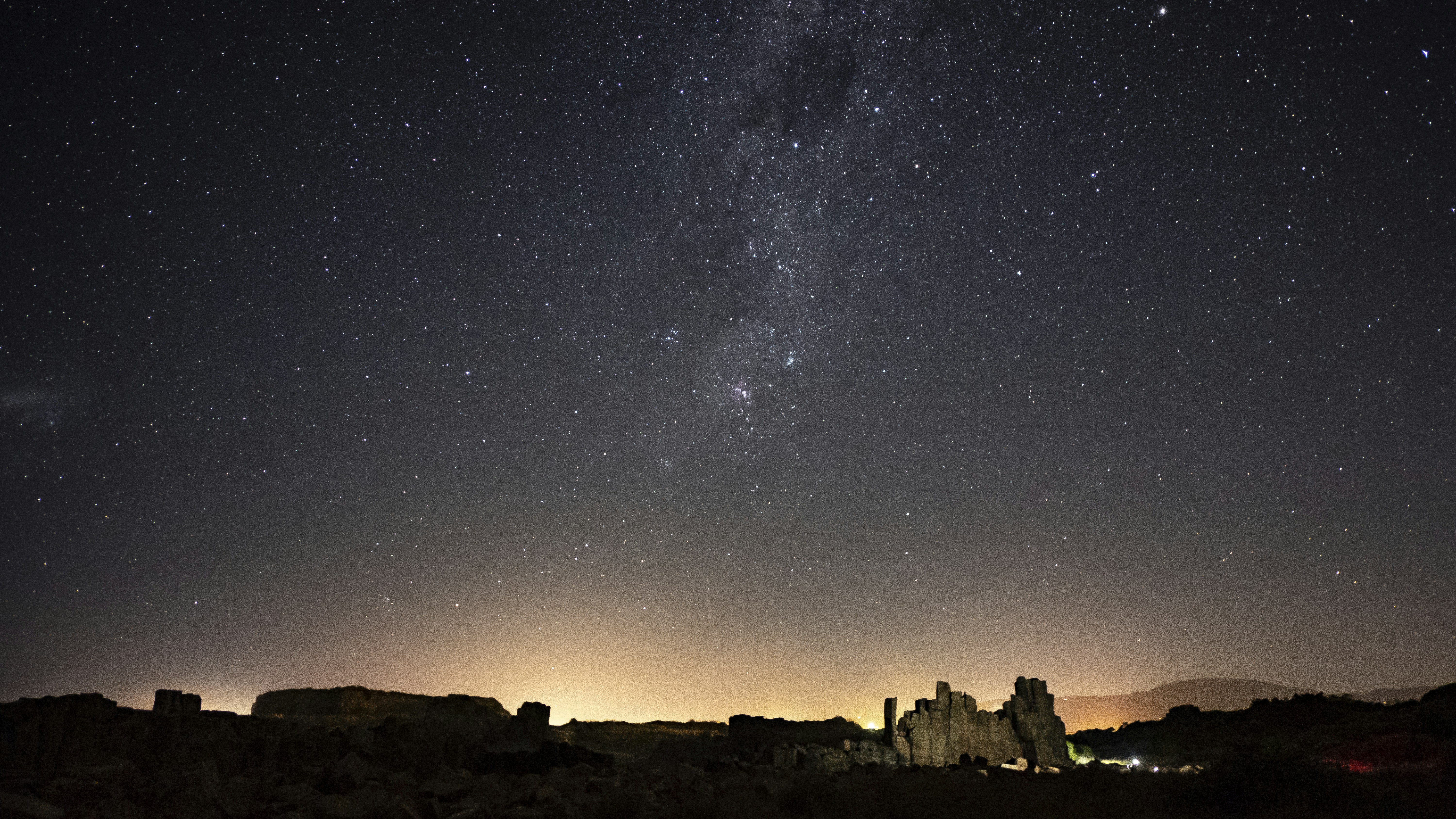 Star-filled sky above rocky desert landscape with distant glow on the horizon.