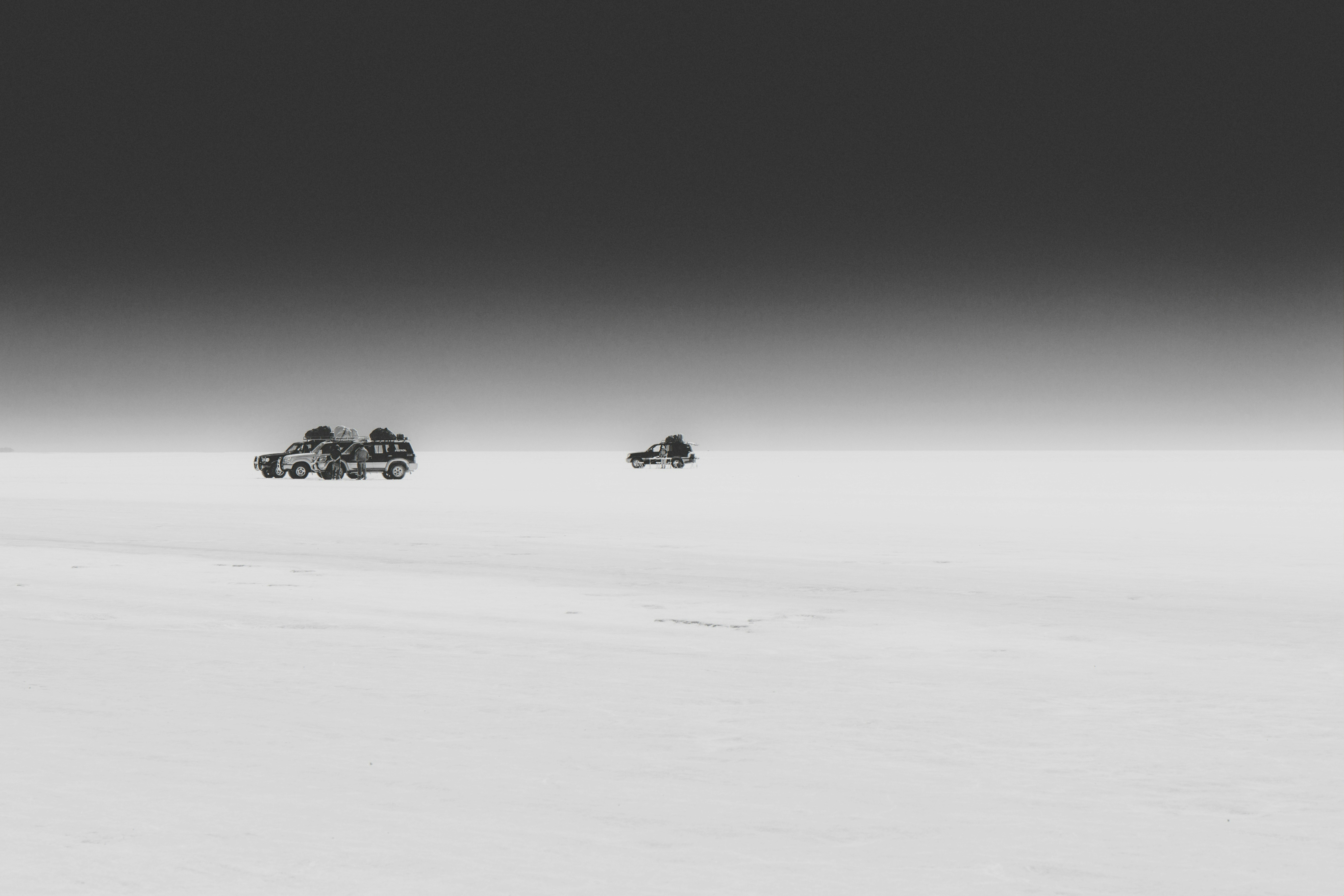 Two vehicles traverse a vast, desolate landscape, surrounded by an expansive, monochromatic horizon. The stark contrast emphasizes the isolation of the scene.
