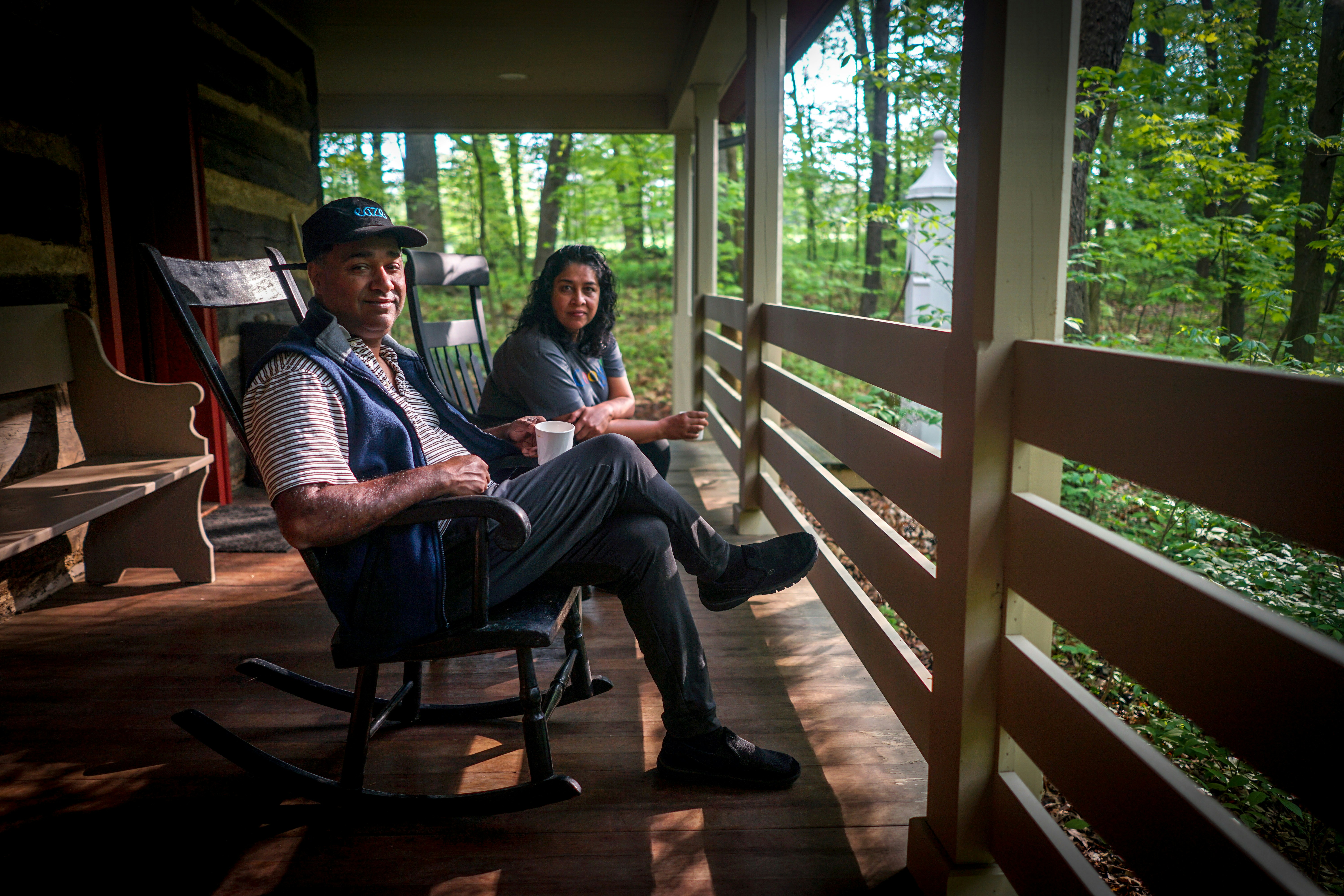 Two people relax on a wooden porch surrounded by lush green forest, with soft sunlight filtering through the trees.