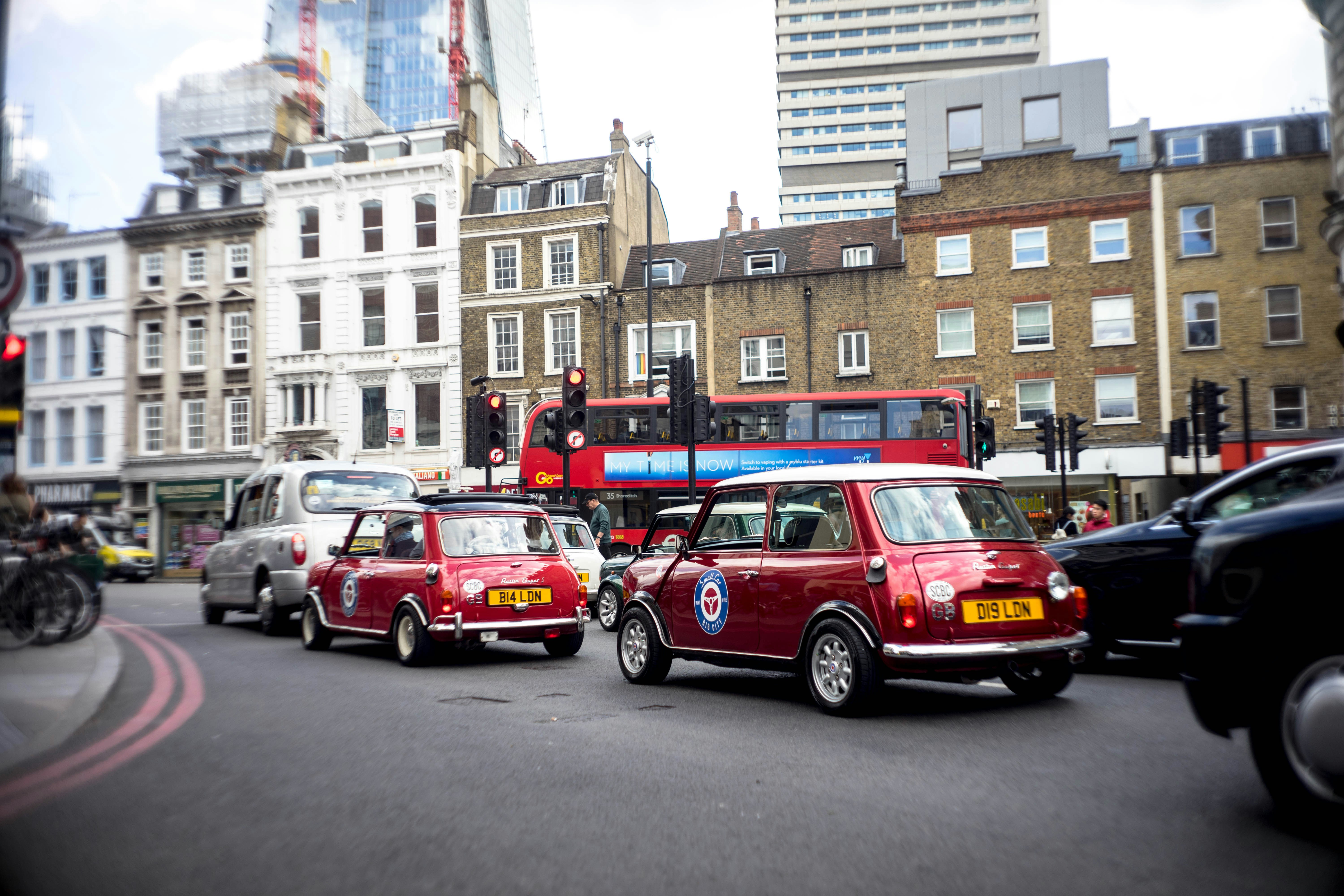 Classic red Mini Coopers driving on a city street lined with historic and modern architecture.