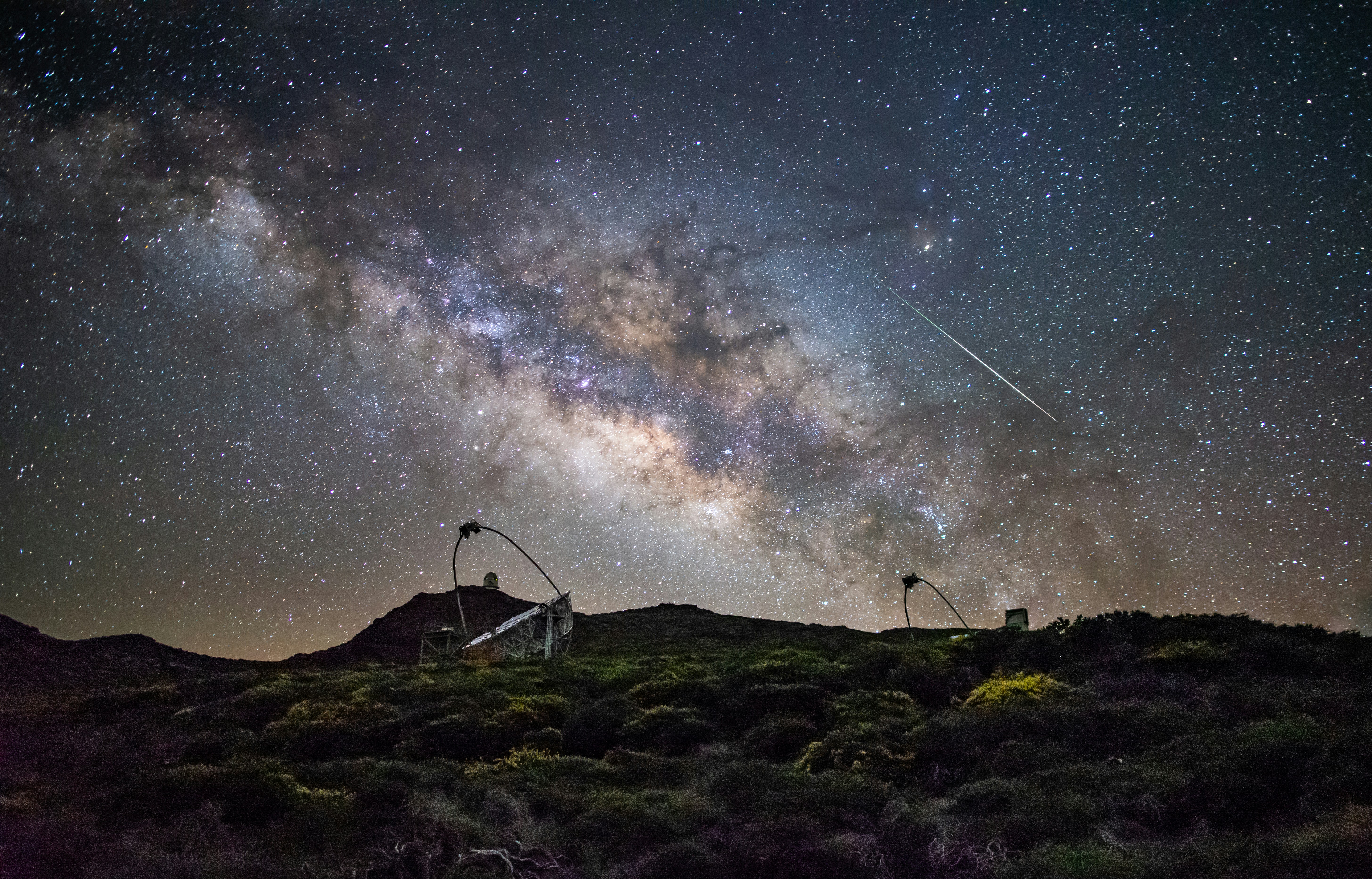 Milky Way galaxy arches over silhouetted telescopes on a rugged landscape under a starry night sky.