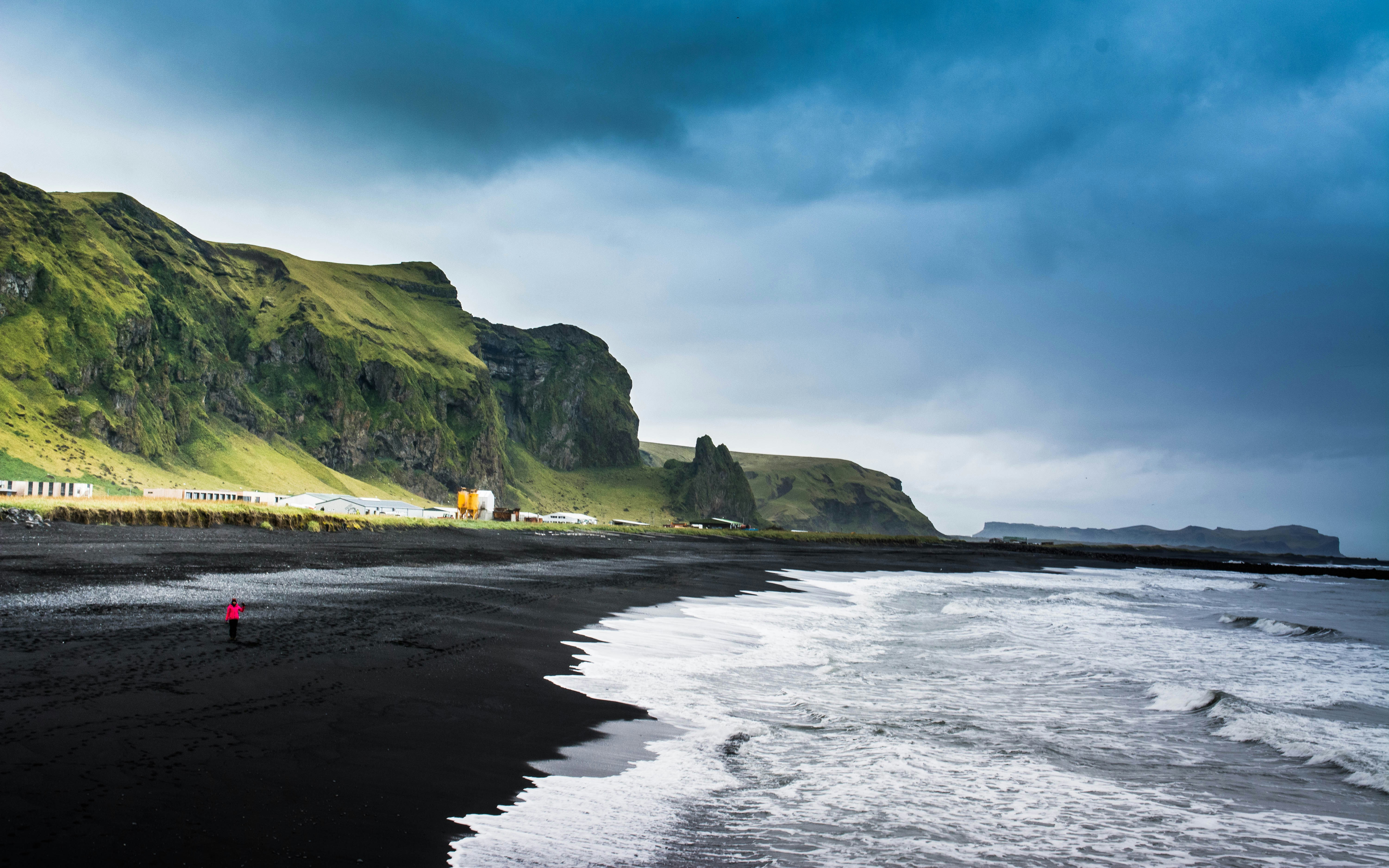 Black sand beach with green cliffs under a cloudy sky, a person in a red jacket walking along the shore.