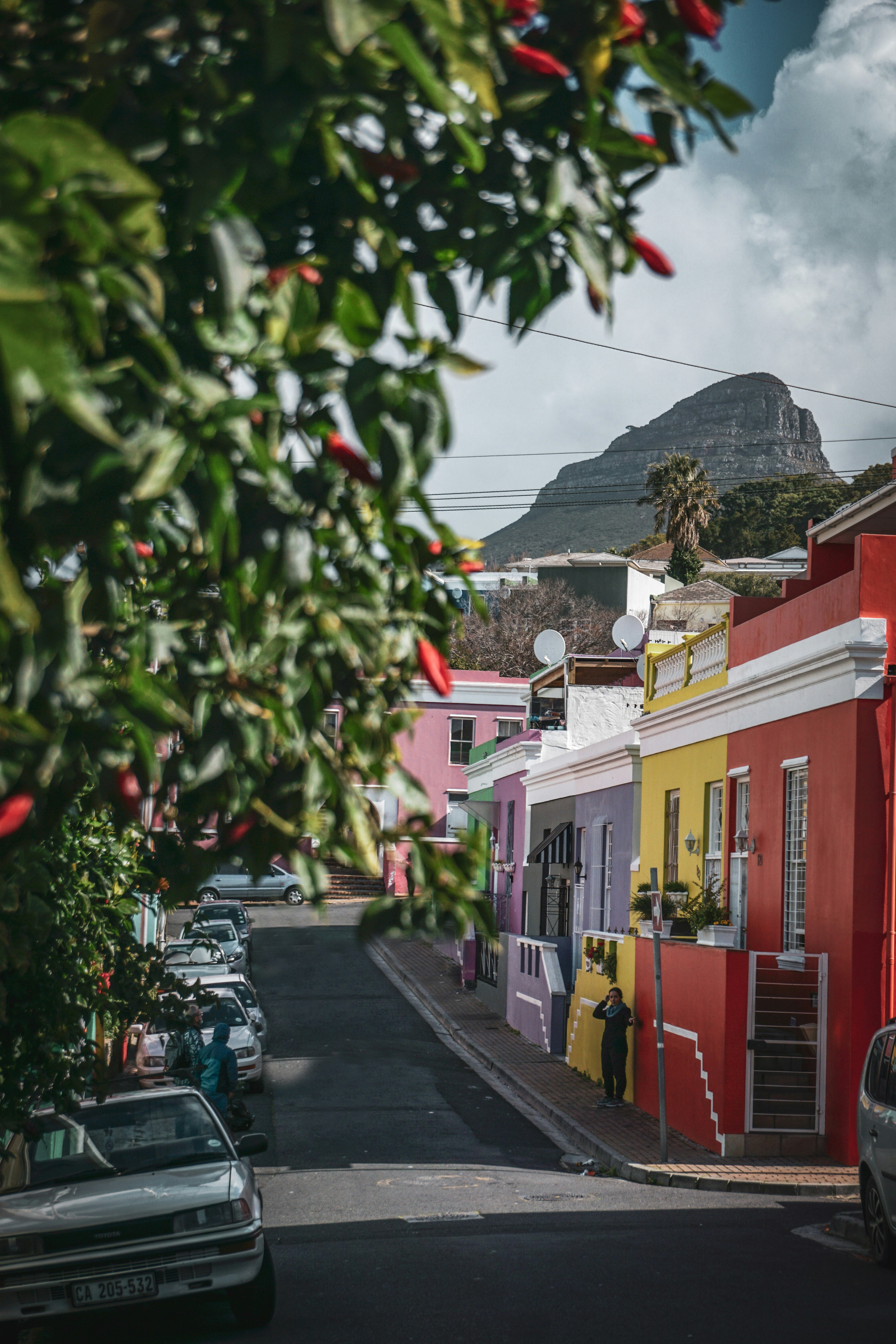 Colorful houses line a street with a mountain backdrop, showcasing the charm of urban life. A figure stands near the vibrant red and yellow buildings.