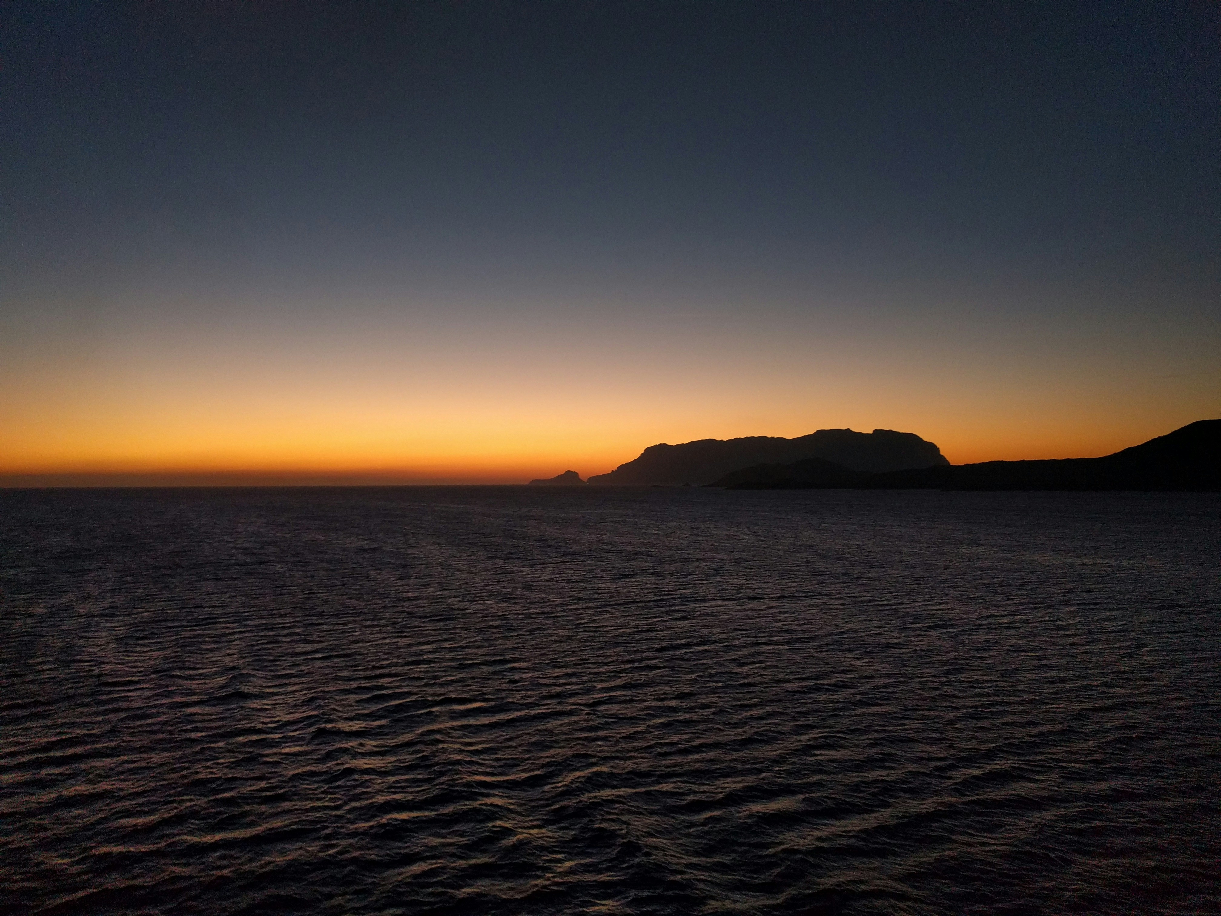 Distant silhouette of Sardinian cliffs against a vibrant sunset sky over a calm sea.