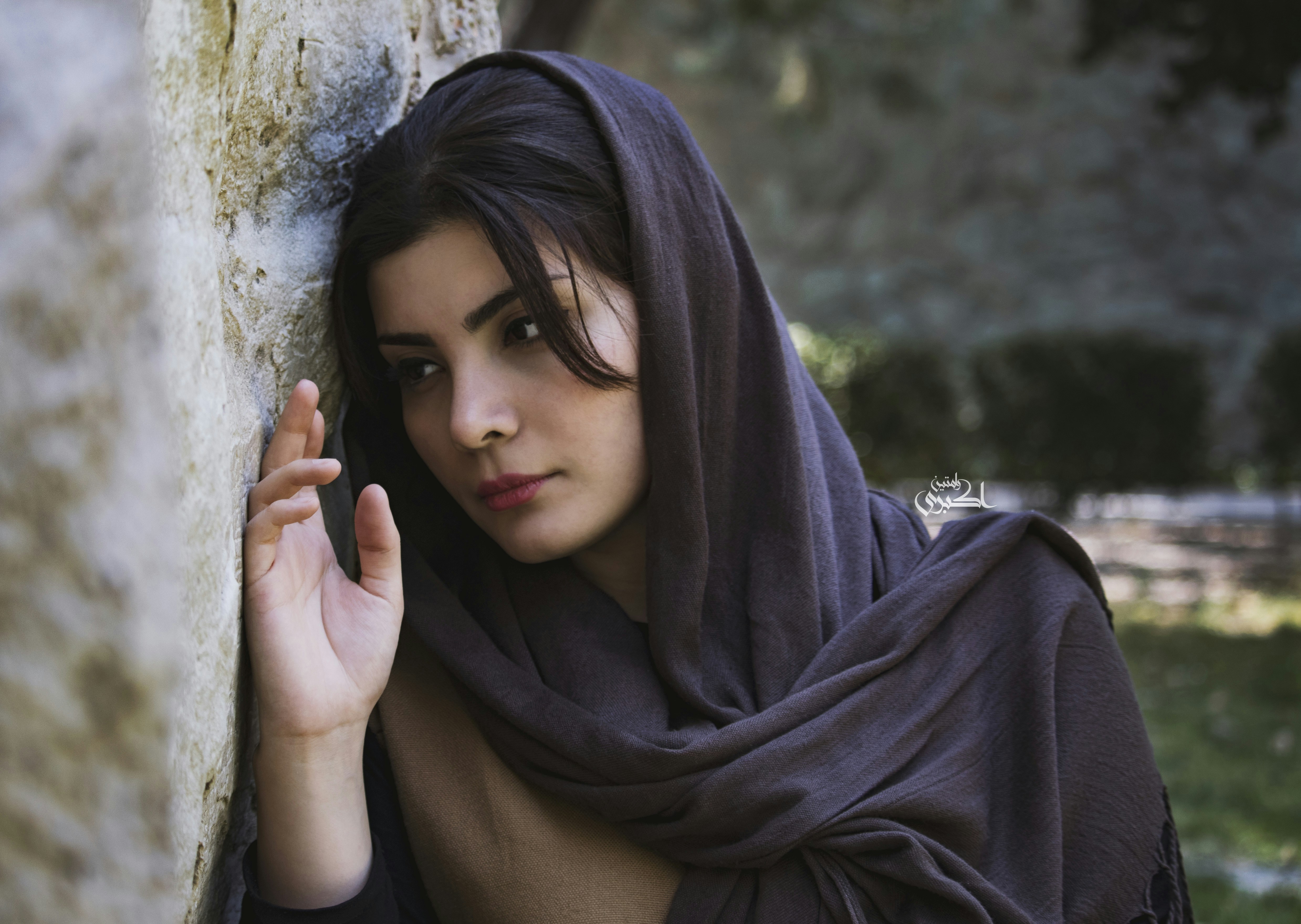A young woman in a brown shawl leans against a textured stone wall, lost in thought amid a serene outdoor setting.