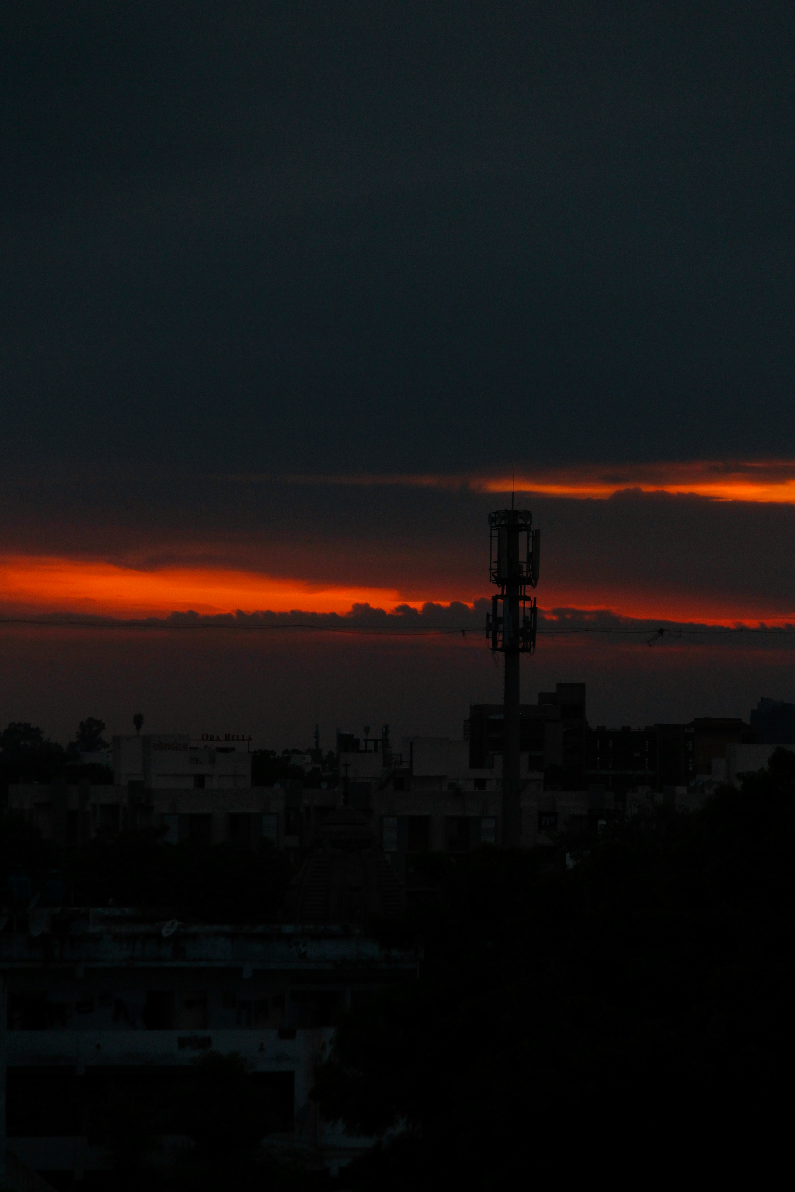 Vibrant orange hues of sunset contrast against dark clouds, highlighting a silhouetted cell tower amidst an urban landscape.