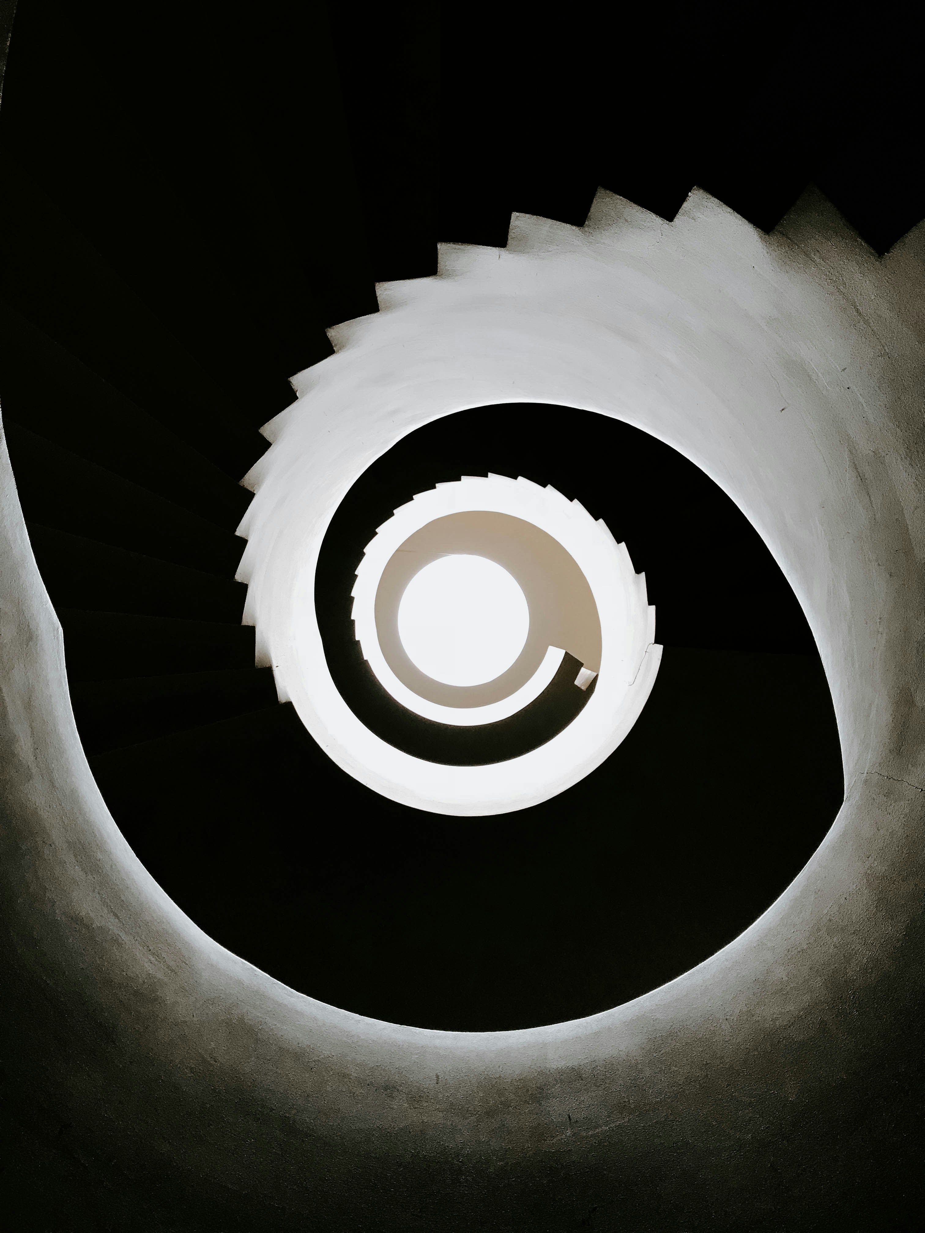 A mesmerizing spiral staircase illuminated by a central light source, creating a dynamic contrast between dark and light. The architectural design draws the eye inward.