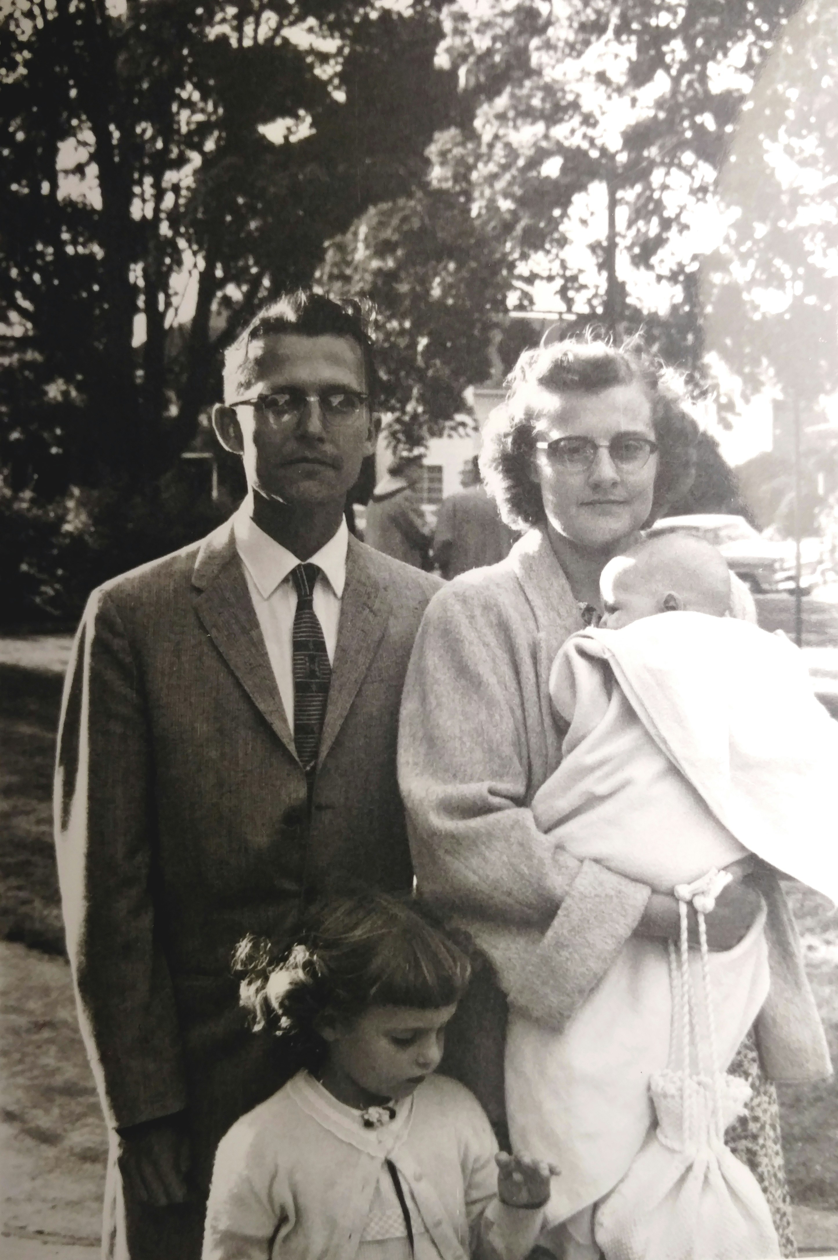 Family of four posing outdoors in black and white, with a man in a suit, a woman holding a baby, and a young girl standing in front.