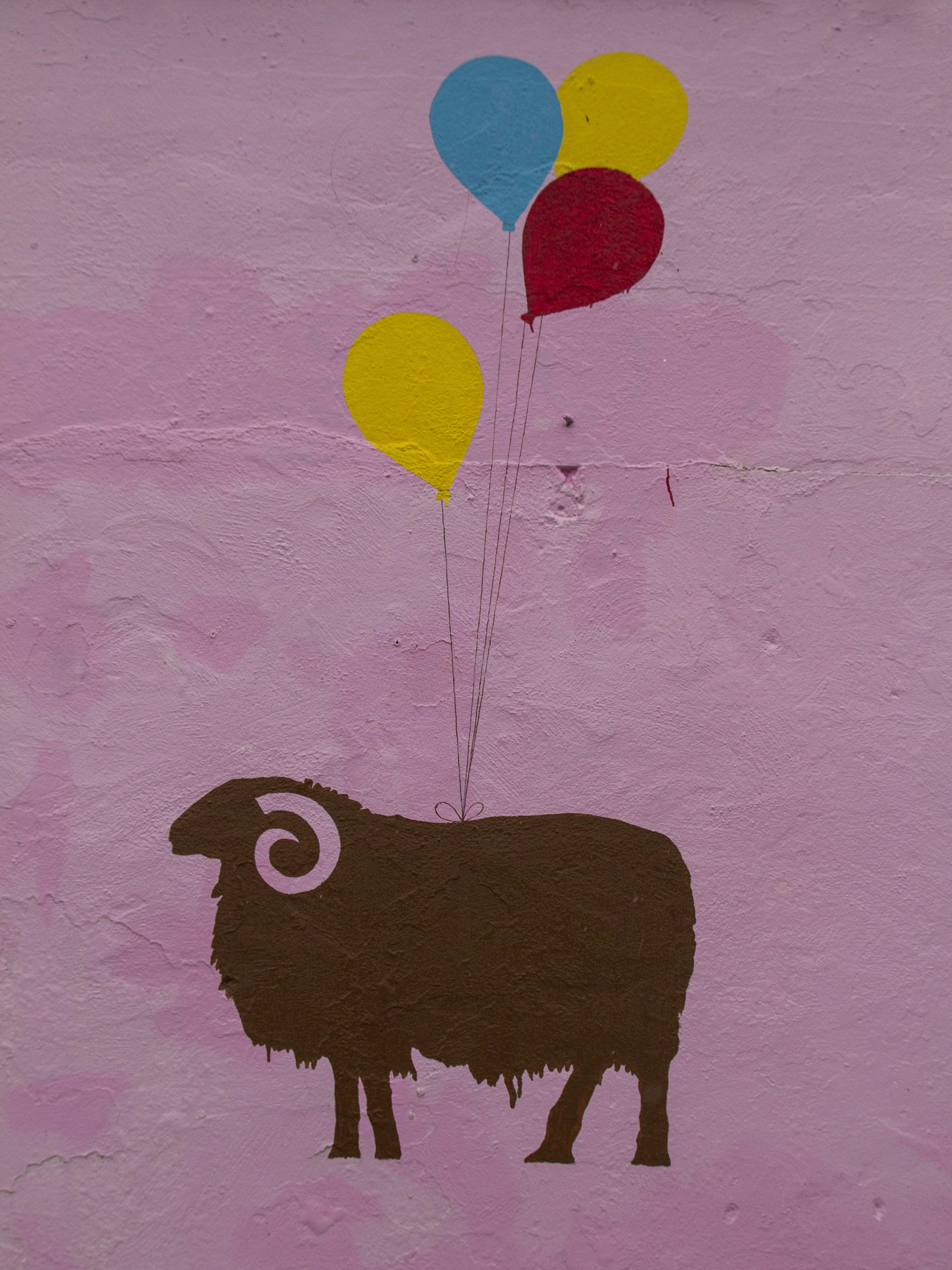 A playful illustration of a brown ram holding three colorful balloons against a pink wall. The image conveys a sense of whimsy and joy.