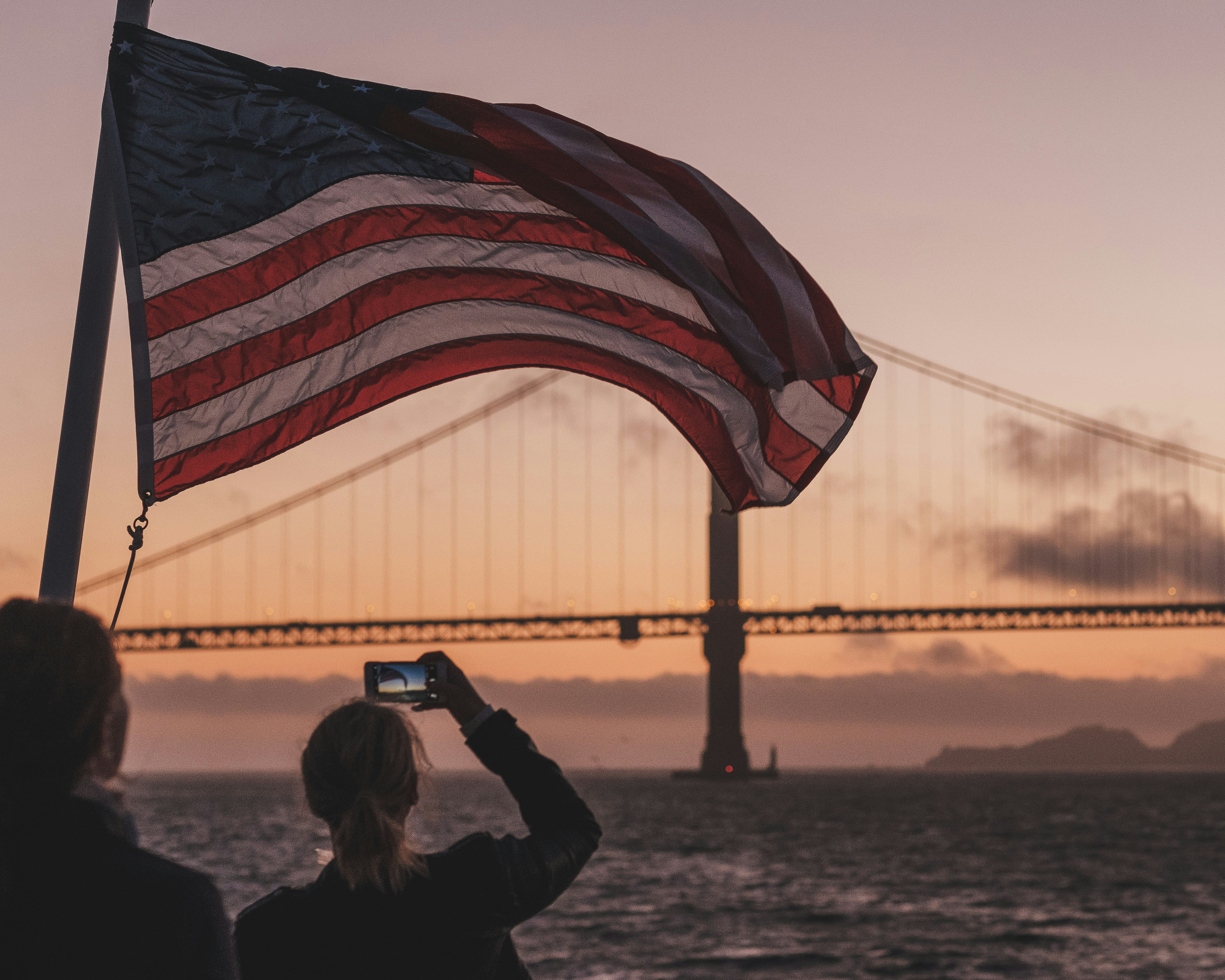 American flag billowing in the breeze as a person captures the Golden Gate Bridge at sunset.