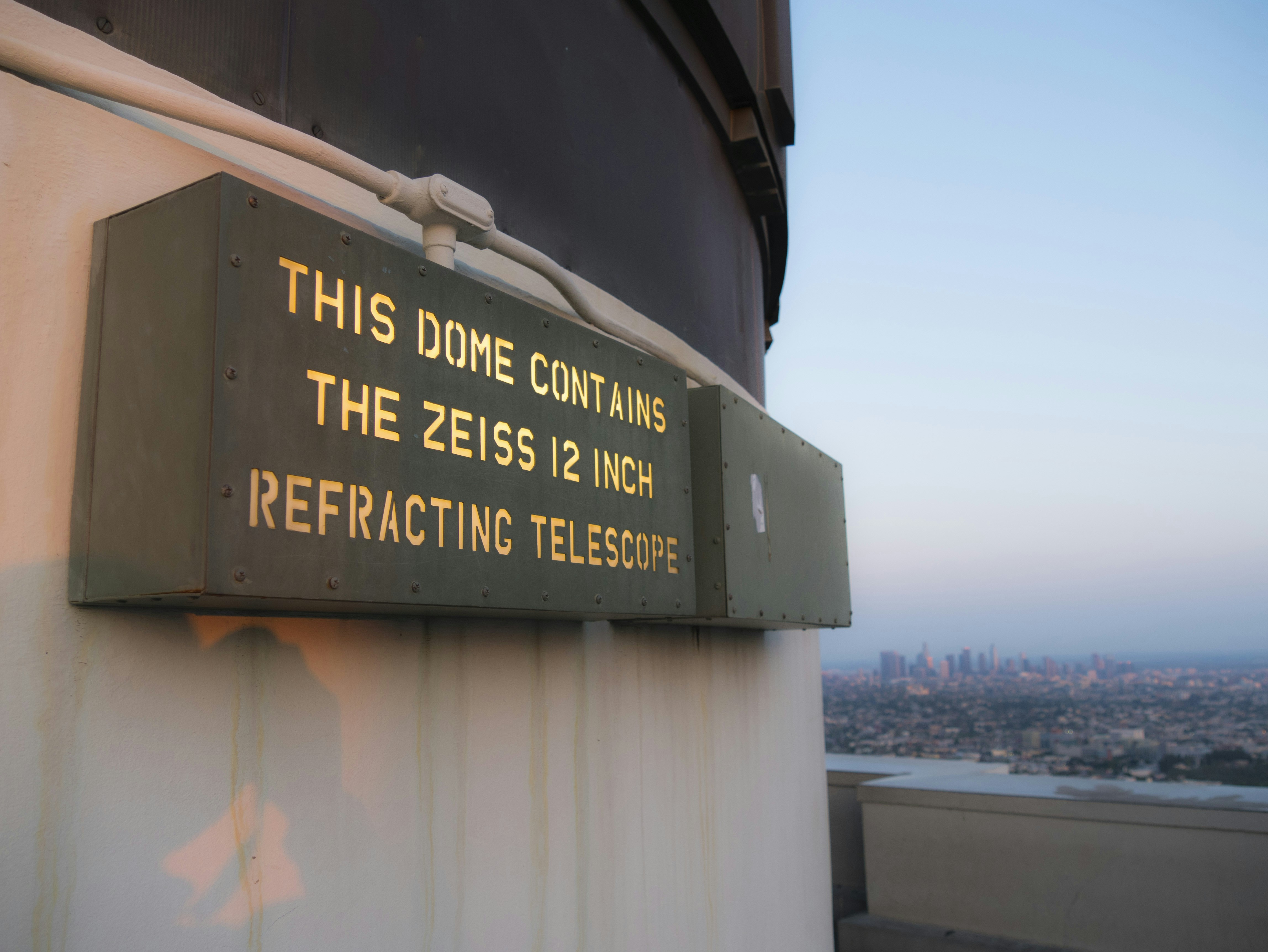 Metal plaque on a dome proclaims a Zeiss 12-inch refracting telescope, with a distant city skyline on the horizon.