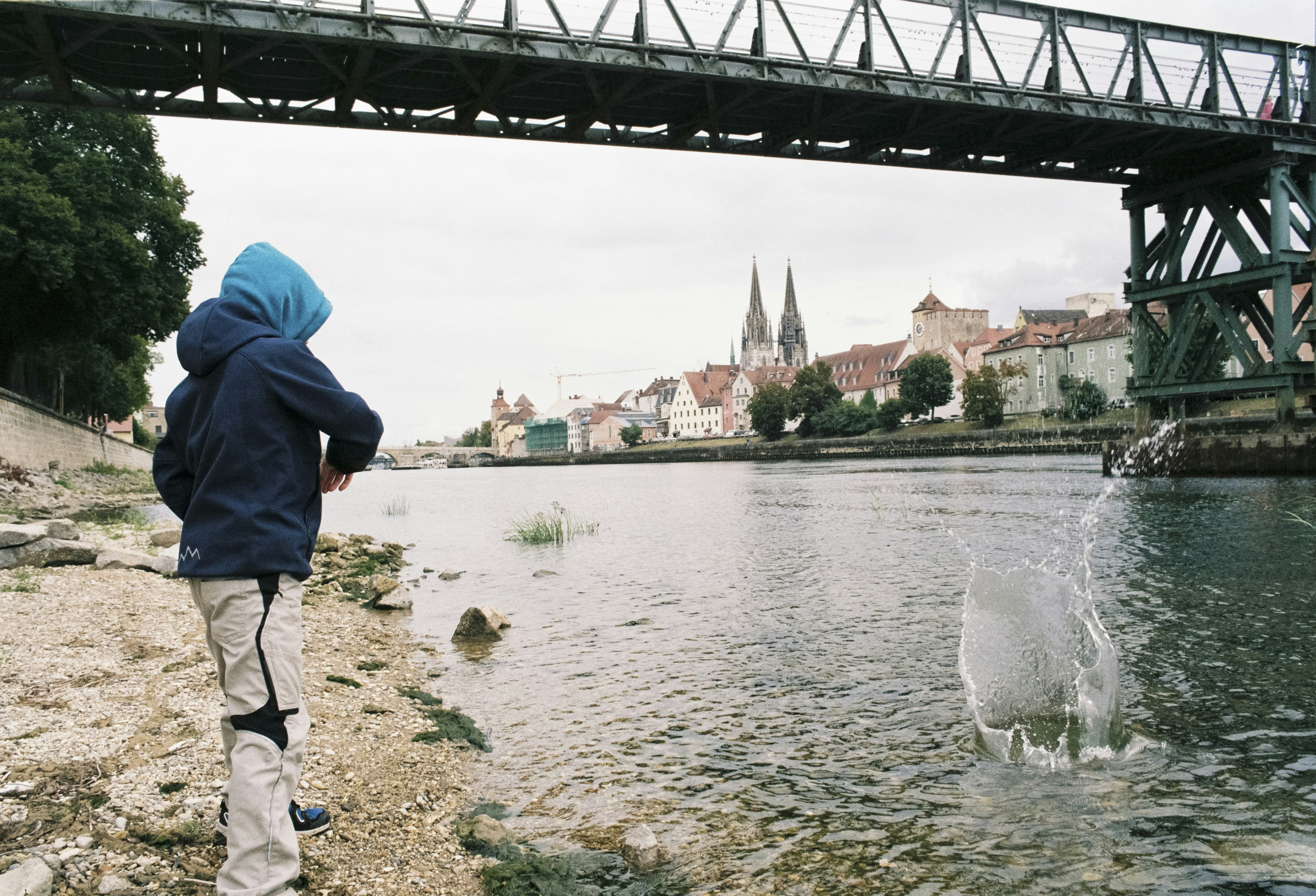 Child in a blue hoodie tossing stones into a river, with a historic cityscape and bridge in the background.