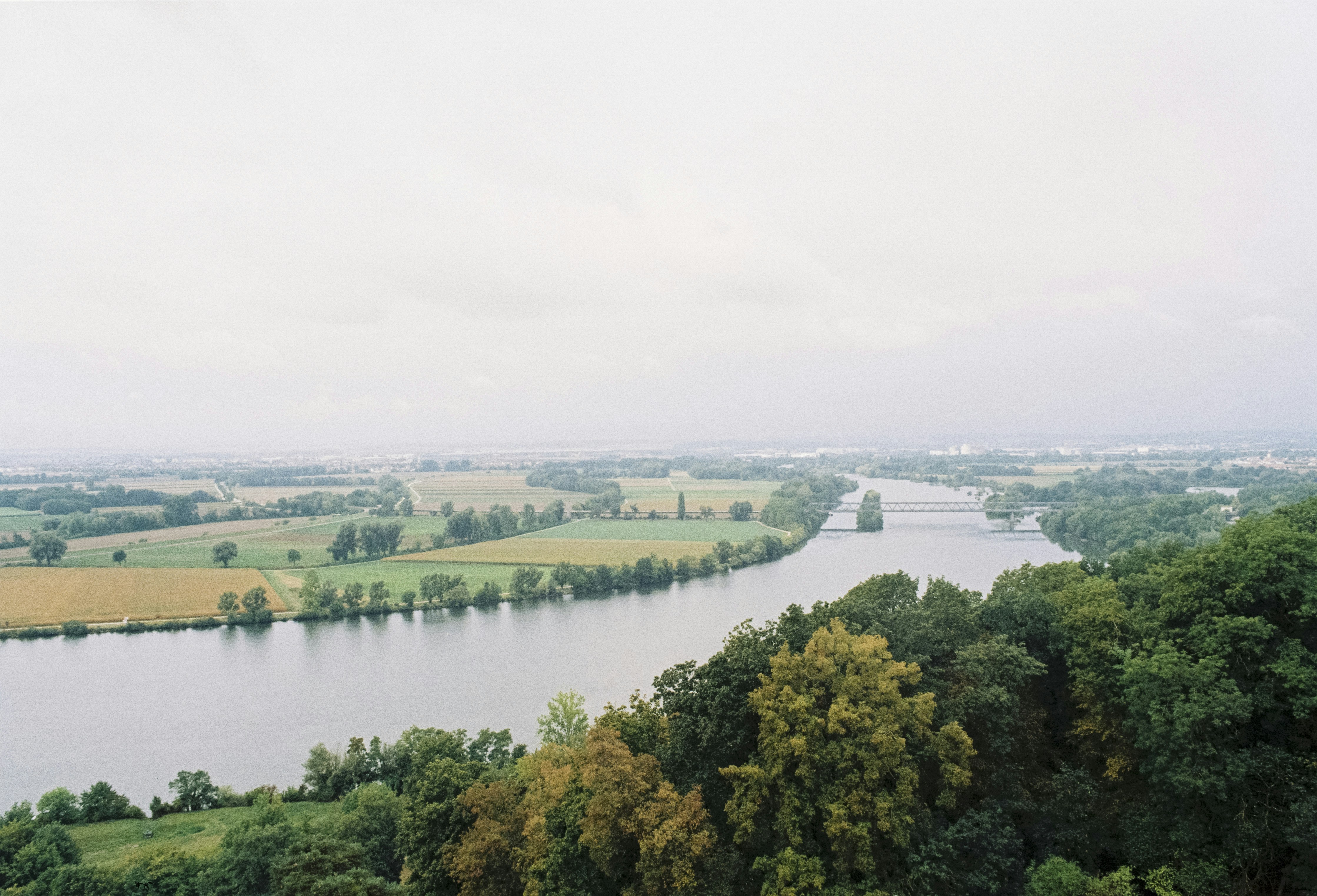 A panoramic view of a winding river bordered by lush greenery and farmland under a cloudy sky.