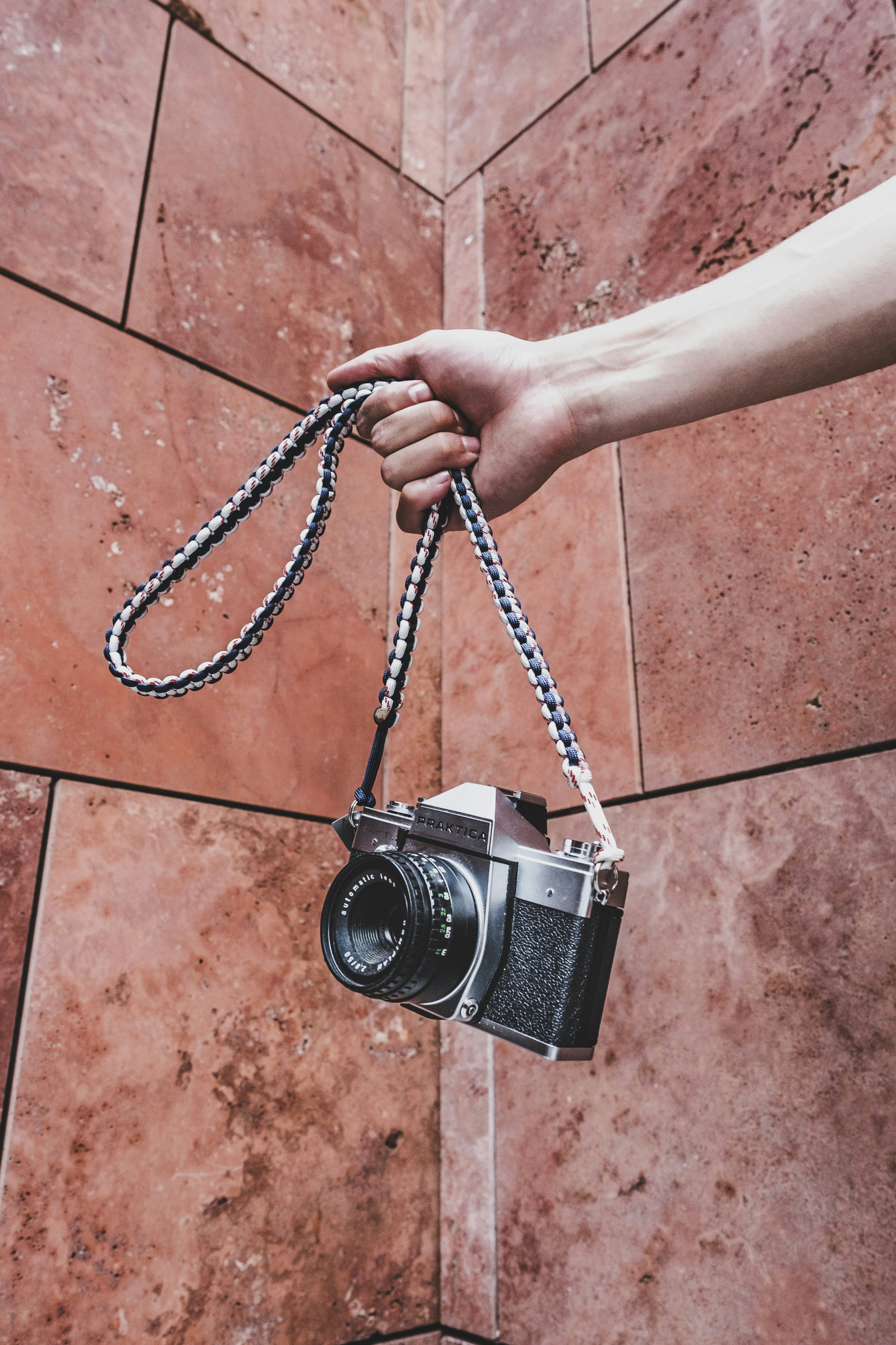 A vintage film camera hangs by a braided strap in front of warm terracotta wall tiles.