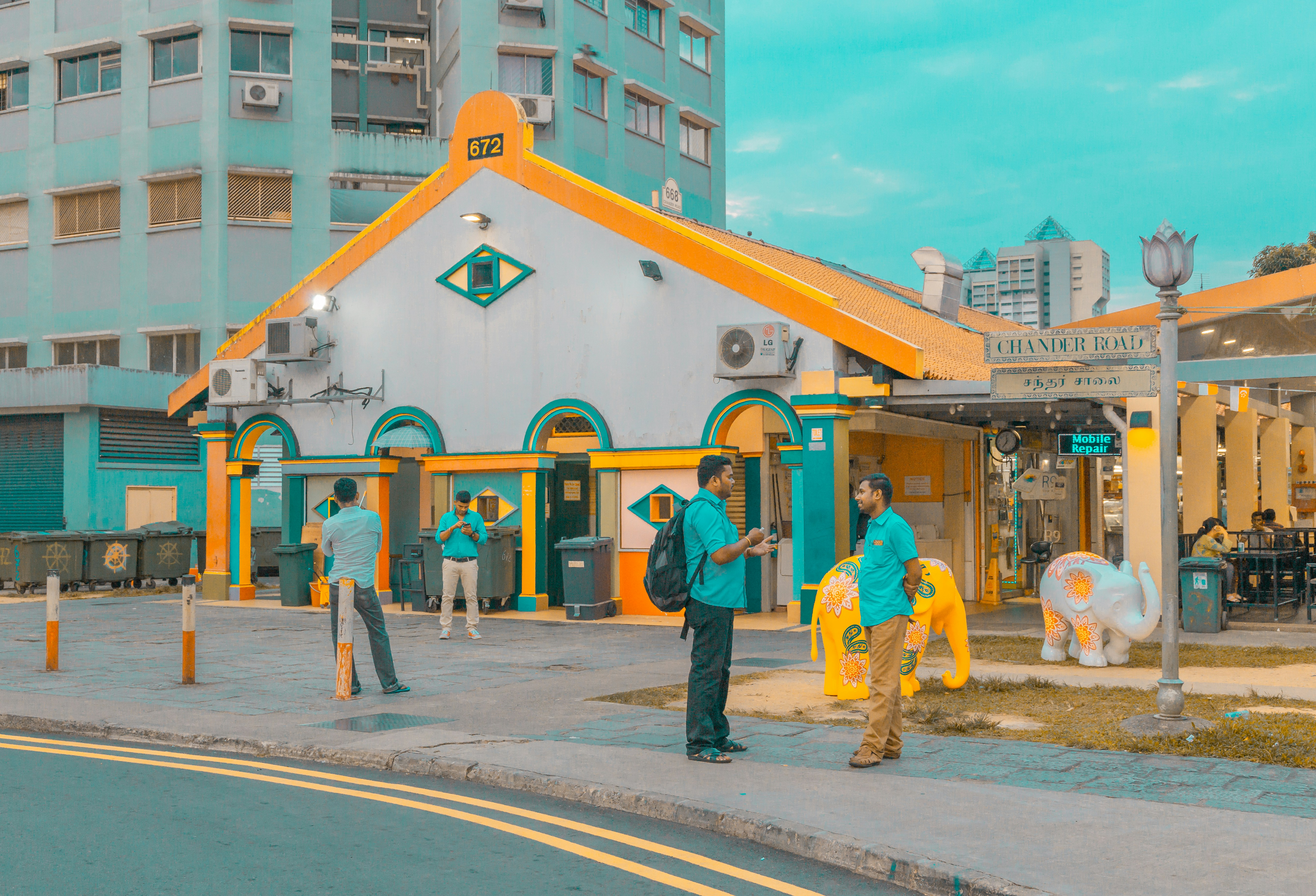 A lively scene featuring individuals in teal attire engaging in conversation outside a brightly painted building with distinctive architectural features.