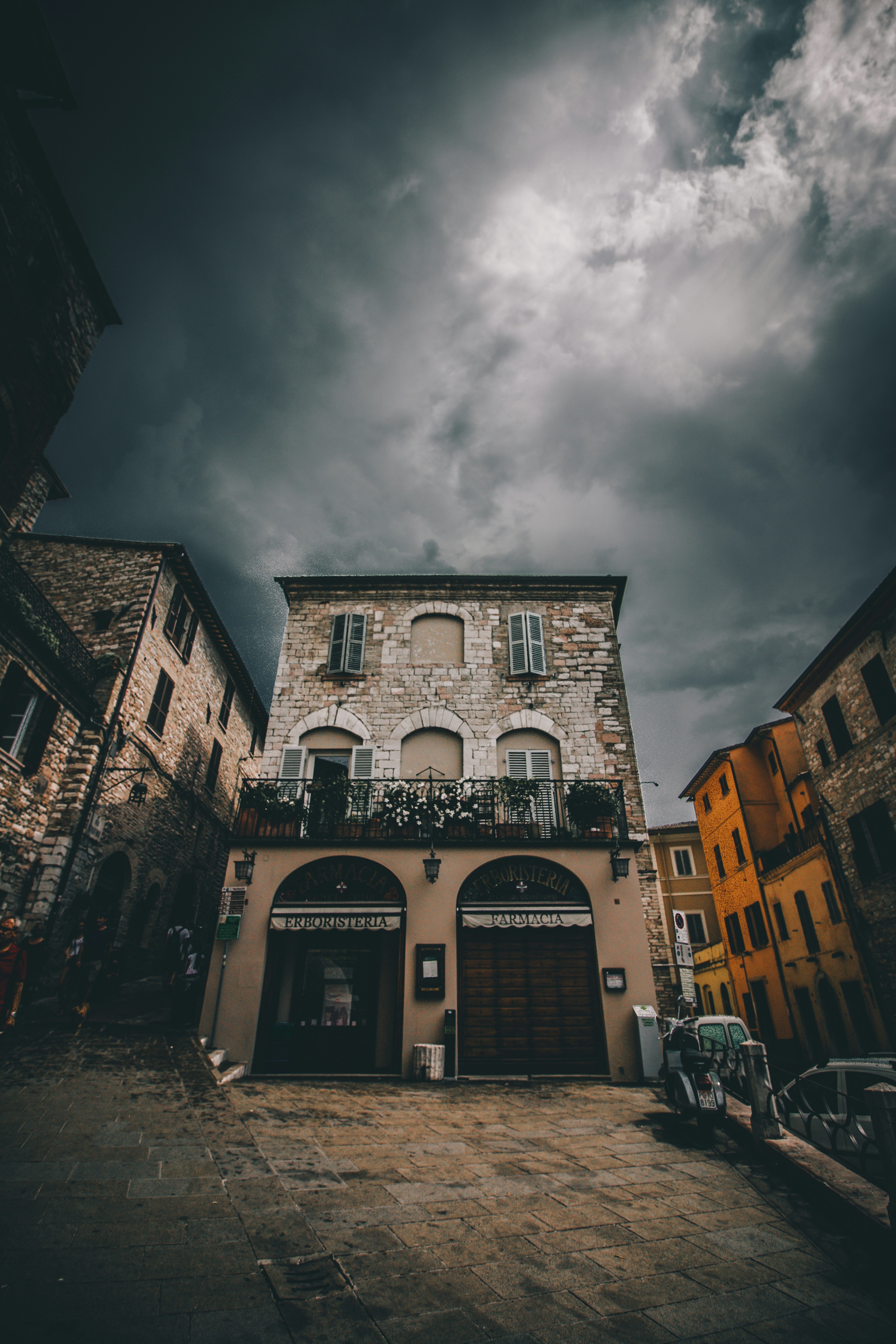 Historic stone building with arched windows and a balcony, surrounded by narrow streets and vibrant facades under a dramatic sky.
