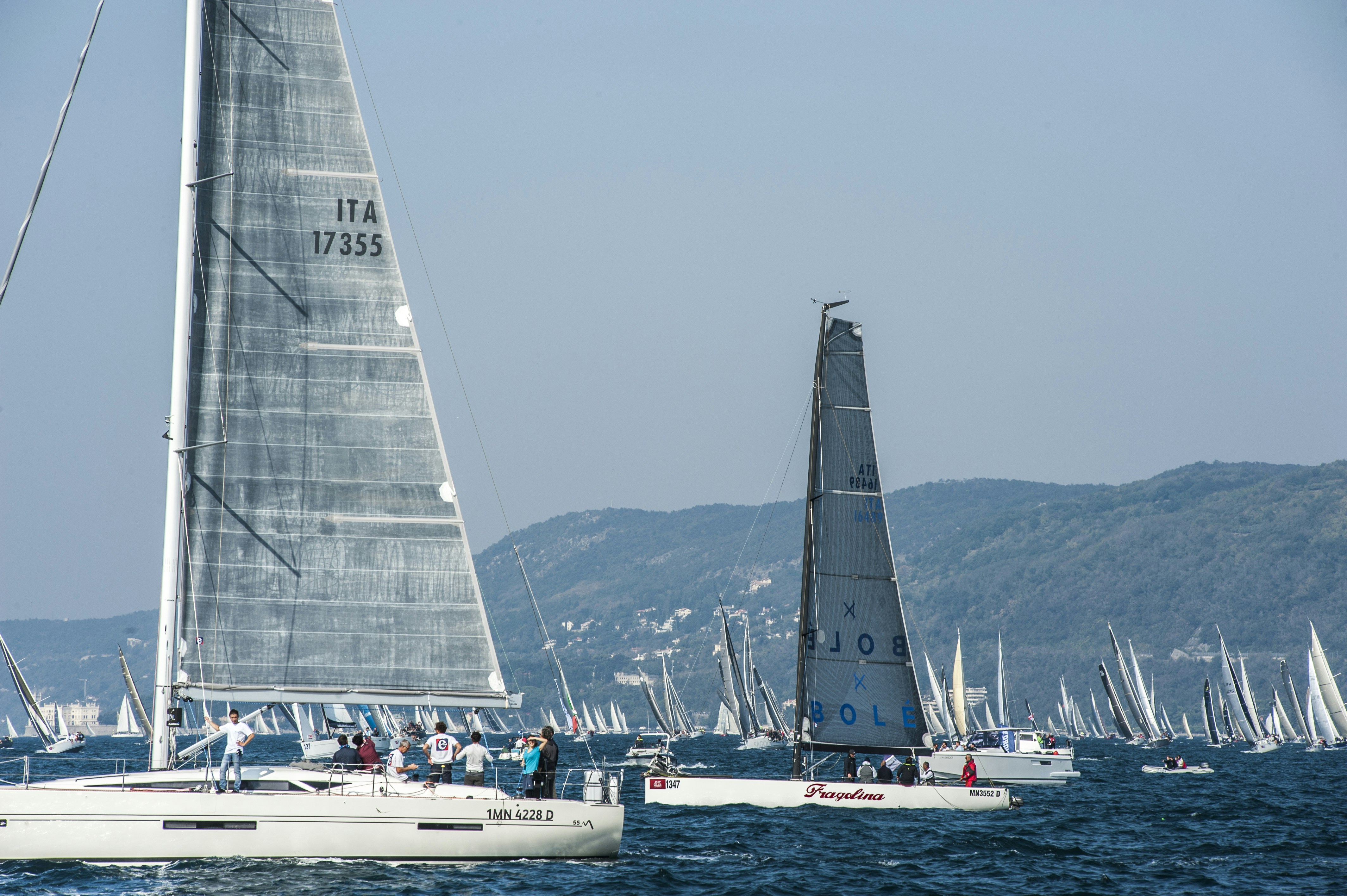 White sailboats glide across a serene body of water with distant hills under a clear sky.