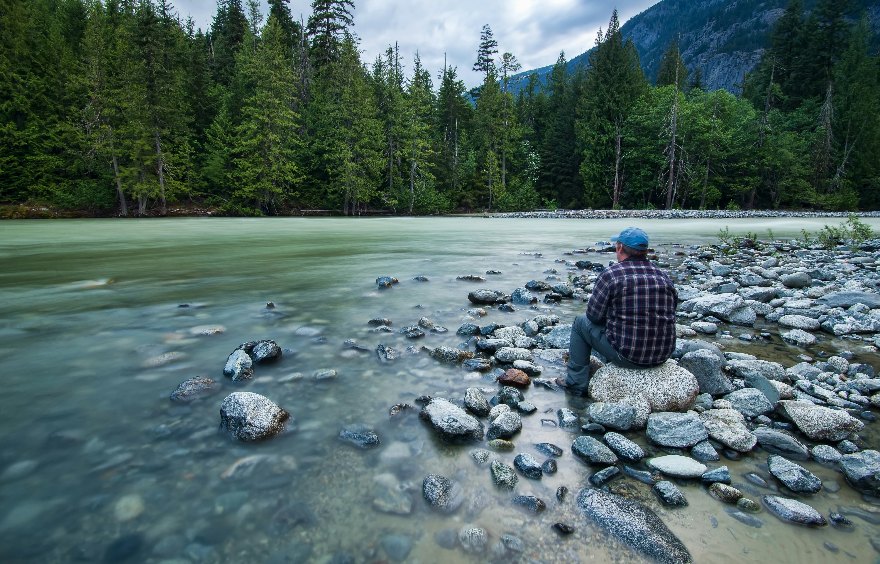 A lone figure sits on a rock by a river, surrounded by lush greenery and mountains, reflecting on the tranquility of nature.