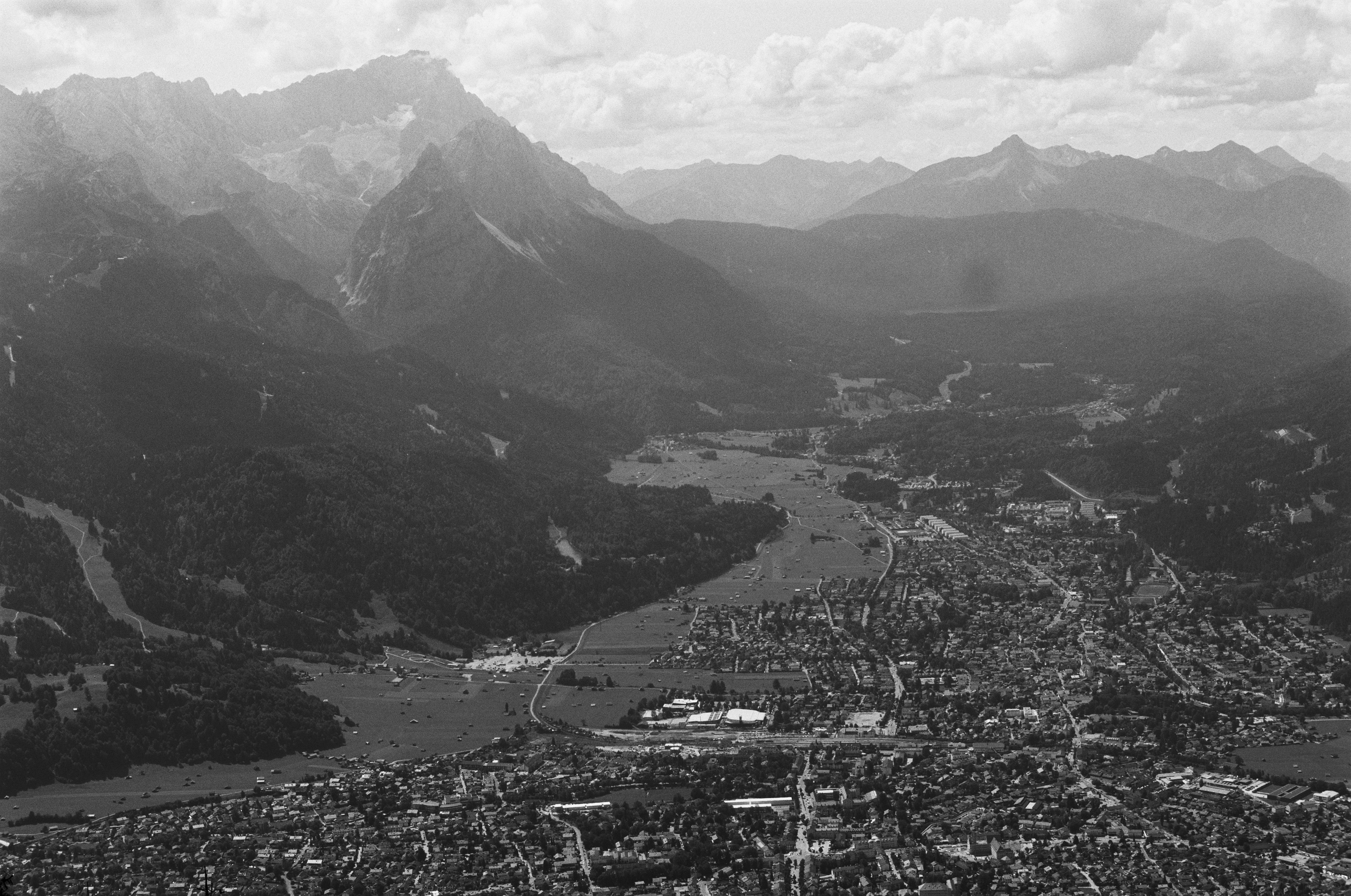 Mountain valley town sprawls beneath towering peaks under a cloudy sky.