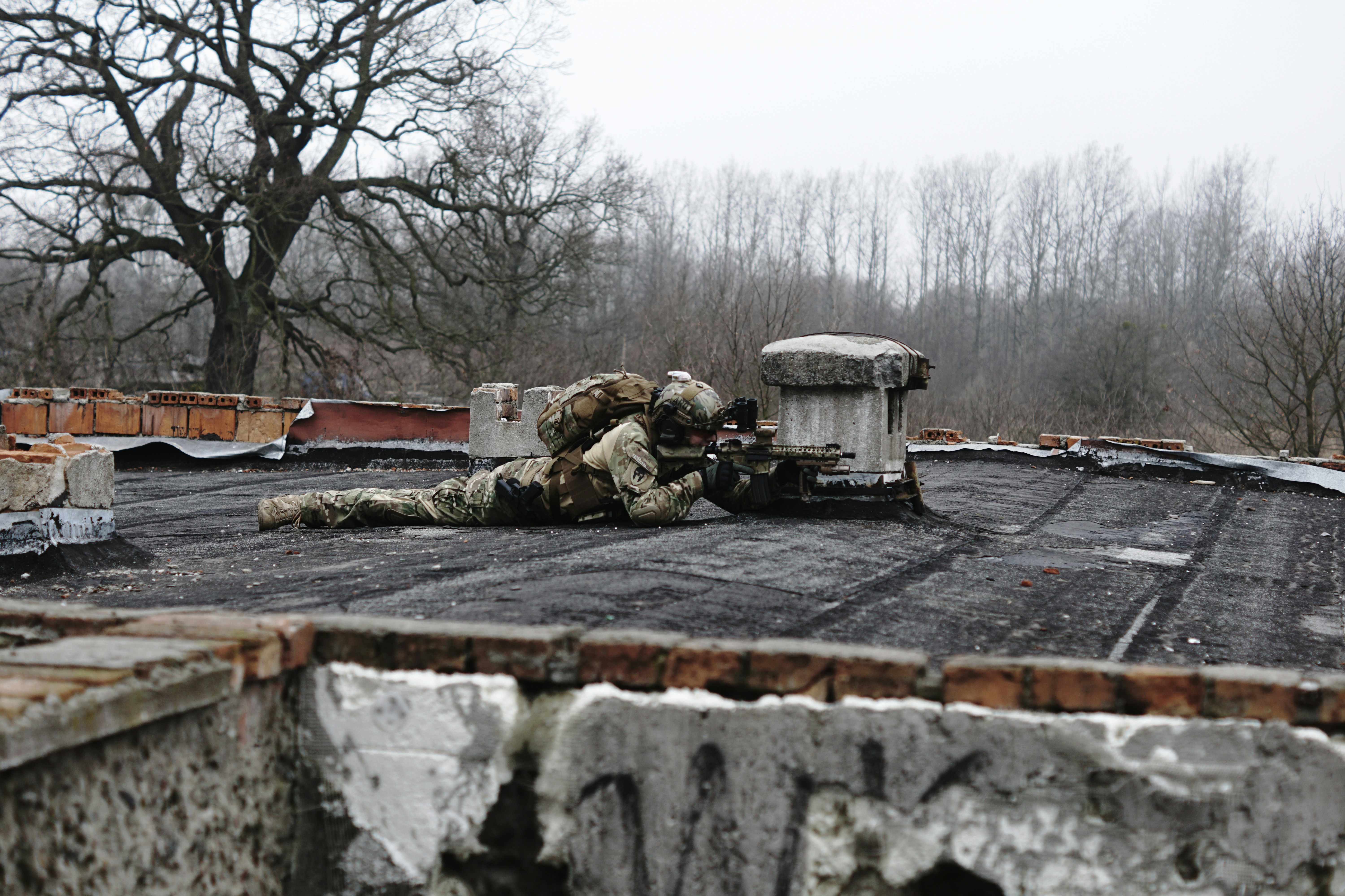 Airsoft player in camouflage gear on a weathered rooftop amid bare winter trees.