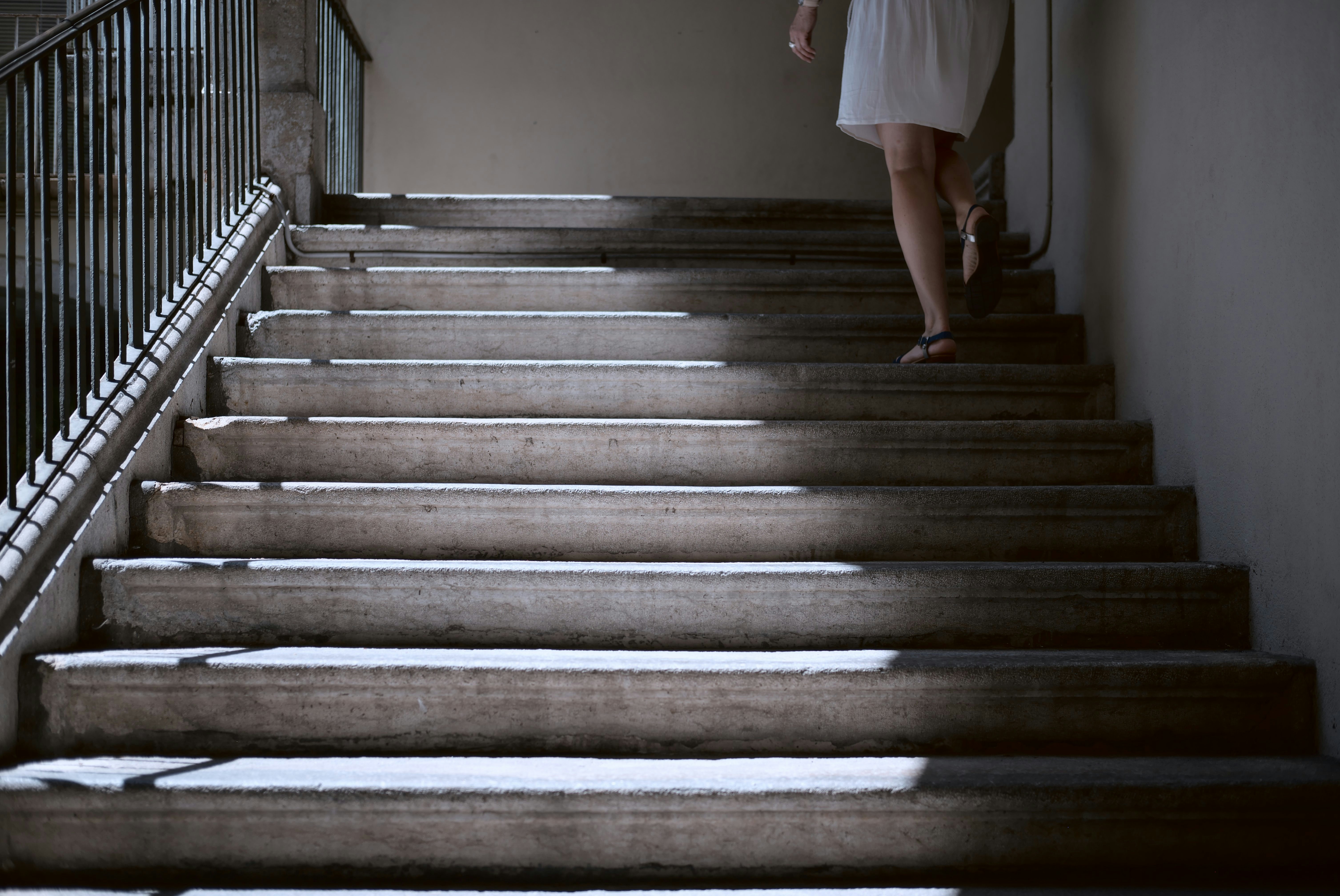 A figure in a white dress ascends a sunlit staircase, casting elongated shadows on the stone steps.