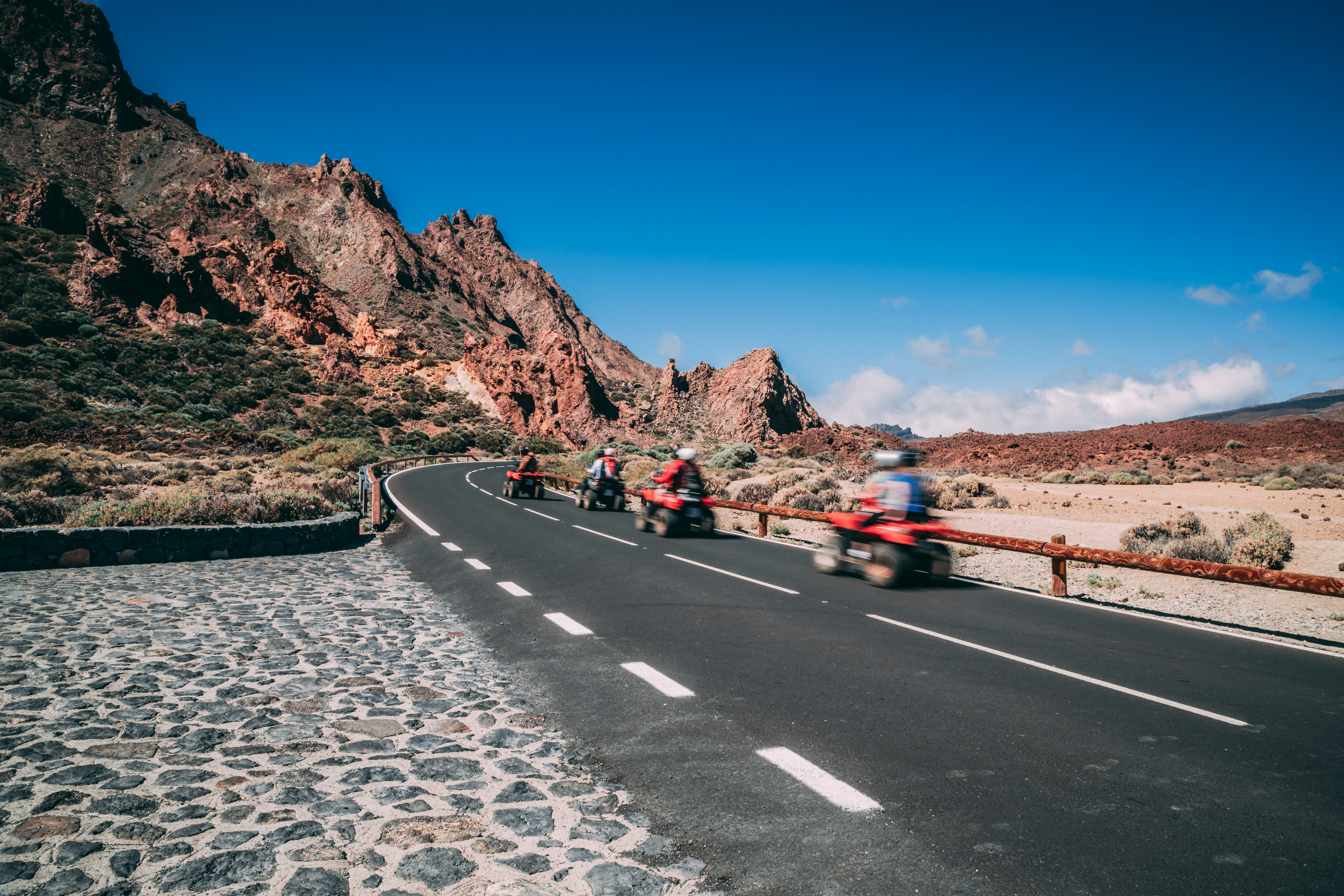 Four people riding ATVs along a winding road through rocky landscape under a clear blue sky.