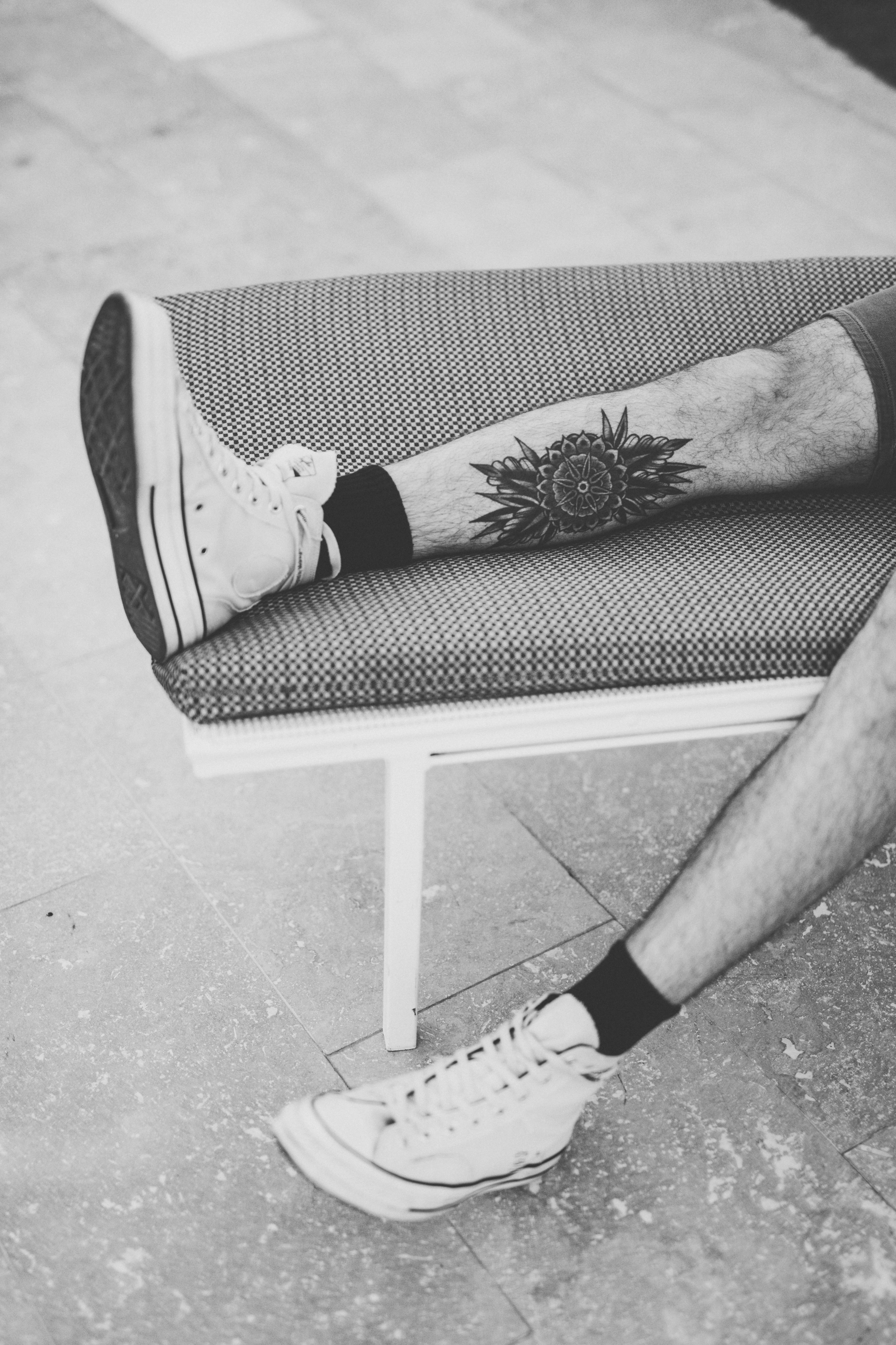 A close-up of a leg adorned with a detailed tattoo, resting on a stylish lounge chair, complemented by classic white sneakers.