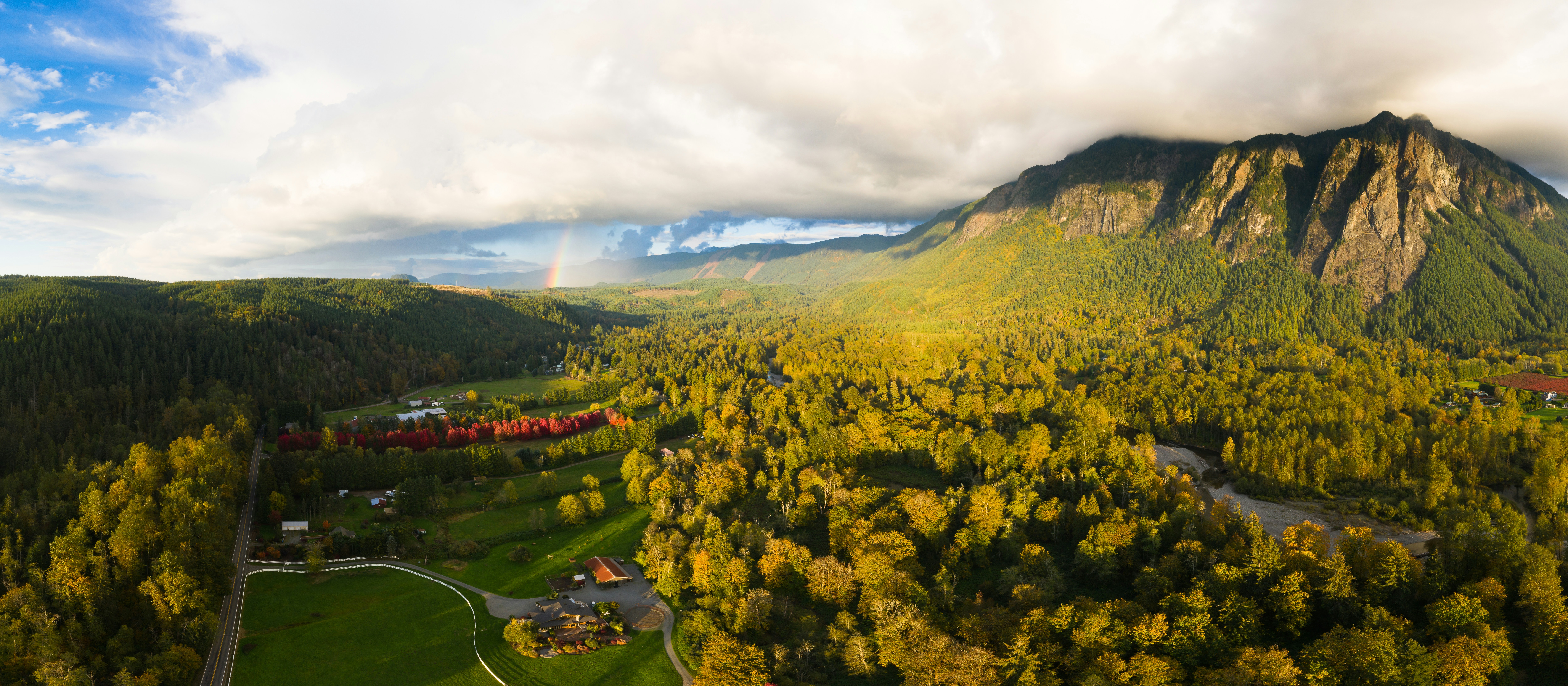 Lush green valley with a winding river and colorful tree line under dramatic clouds and a distant rainbow.