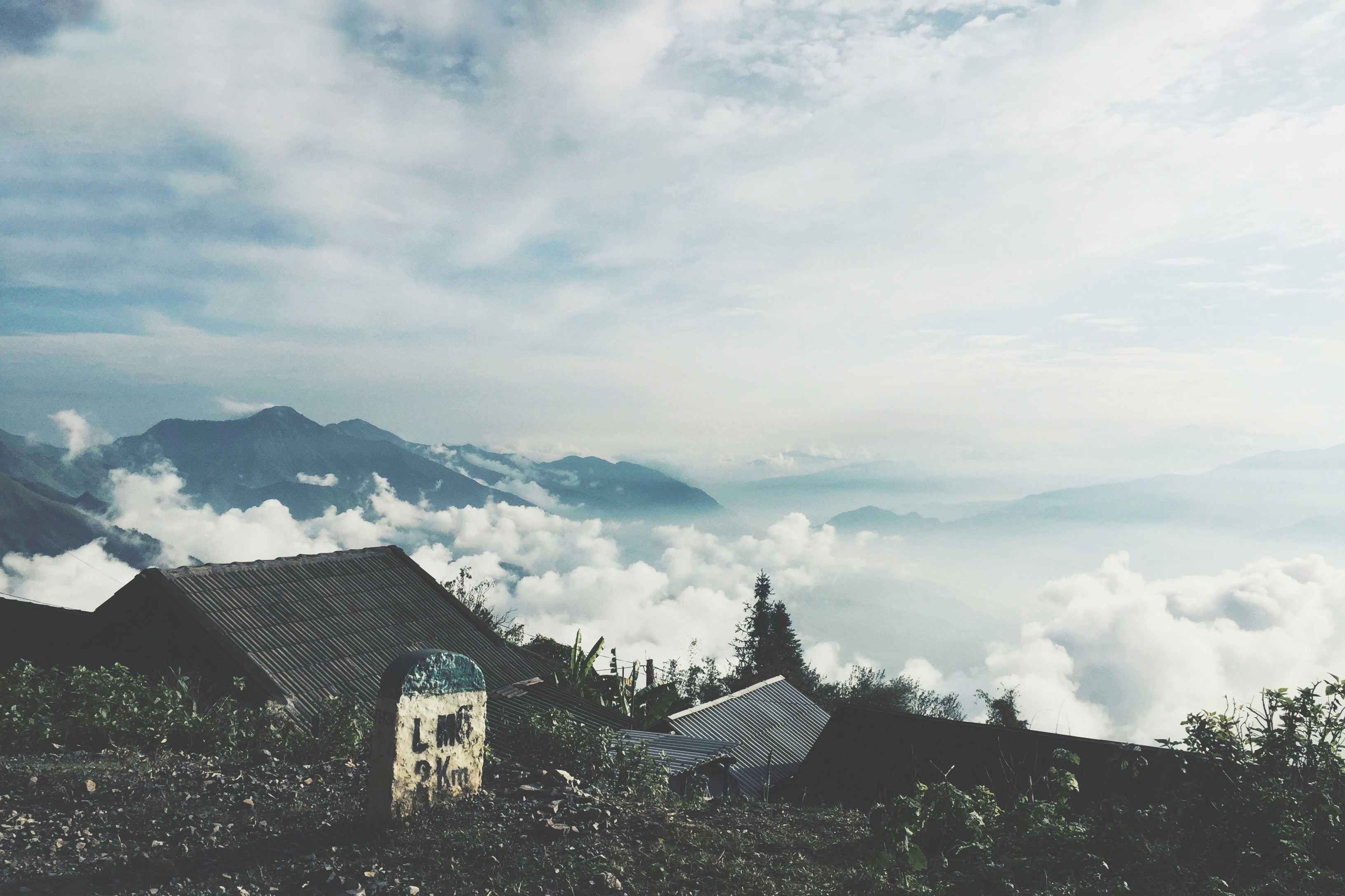 A weathered milestone stands on a mountain ridge, overlooking a sea of clouds and distant peaks under a soft, cloudy sky.