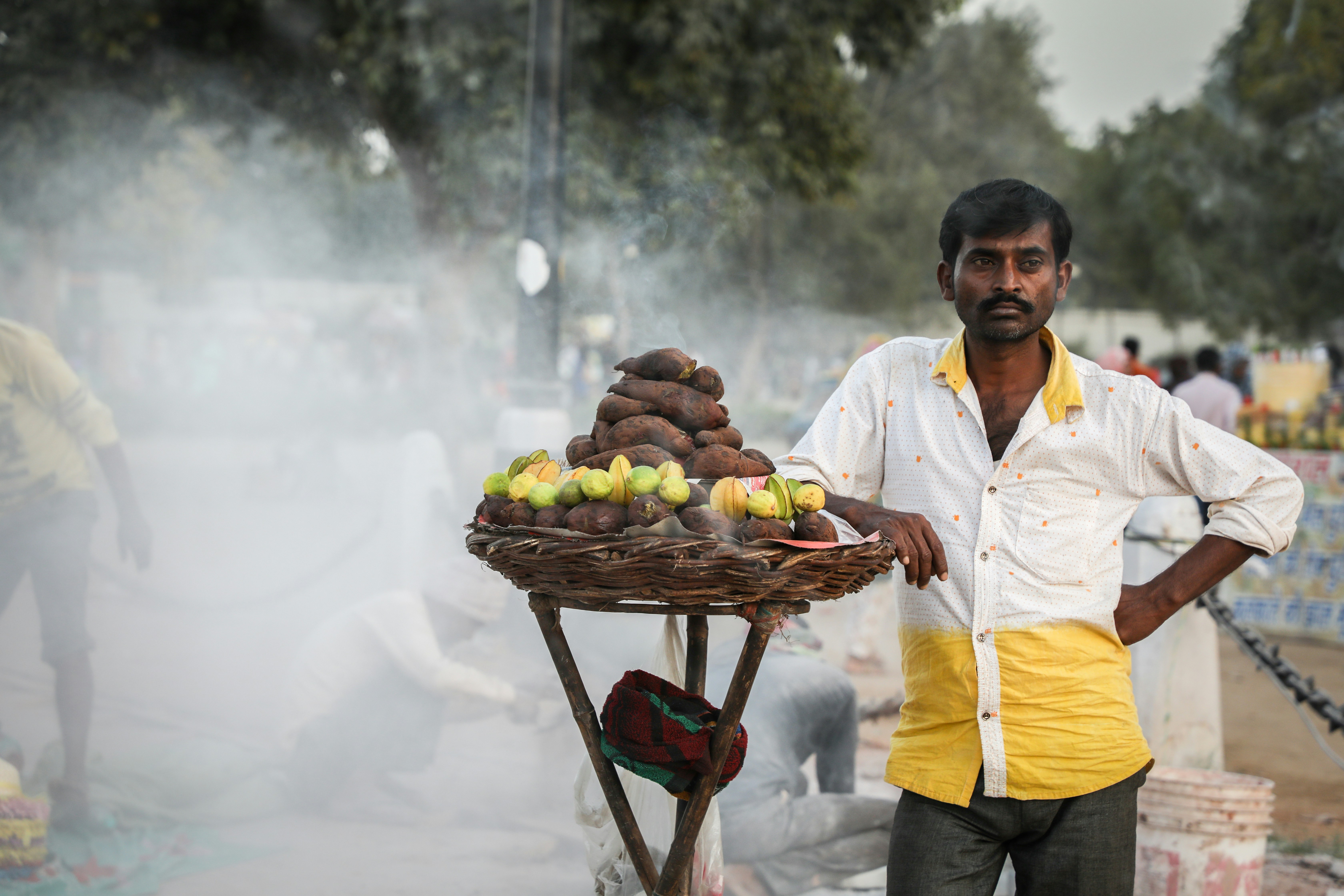 Street vendor stands next to a basket of fresh produce, surrounded by a misty atmosphere in a bustling market scene.