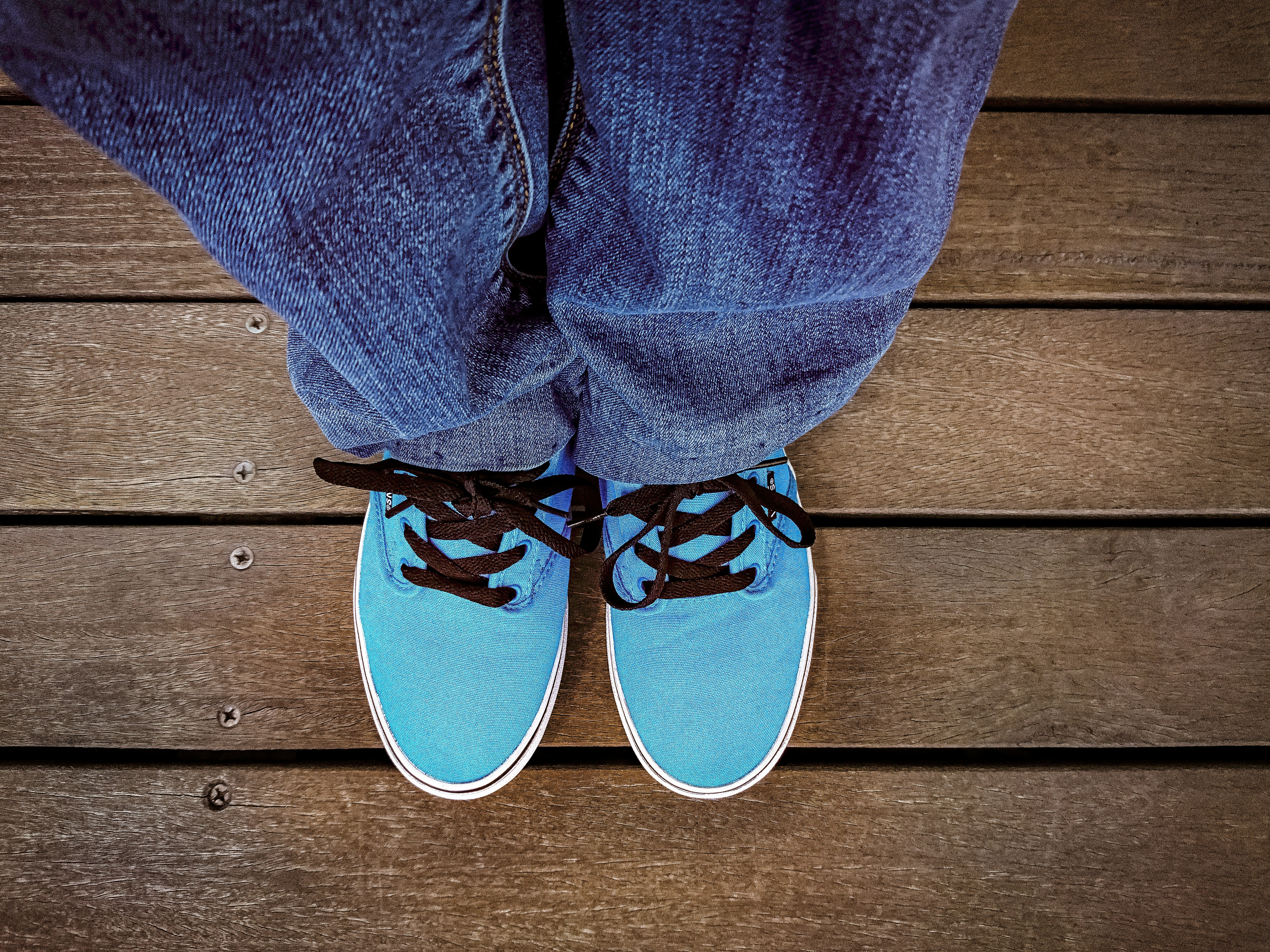 Blue sneakers with brown laces photographed from above on wooden planks, emphasizing casual footwear. The image is a simple lifestyle shot featuring the lower legs and feet.