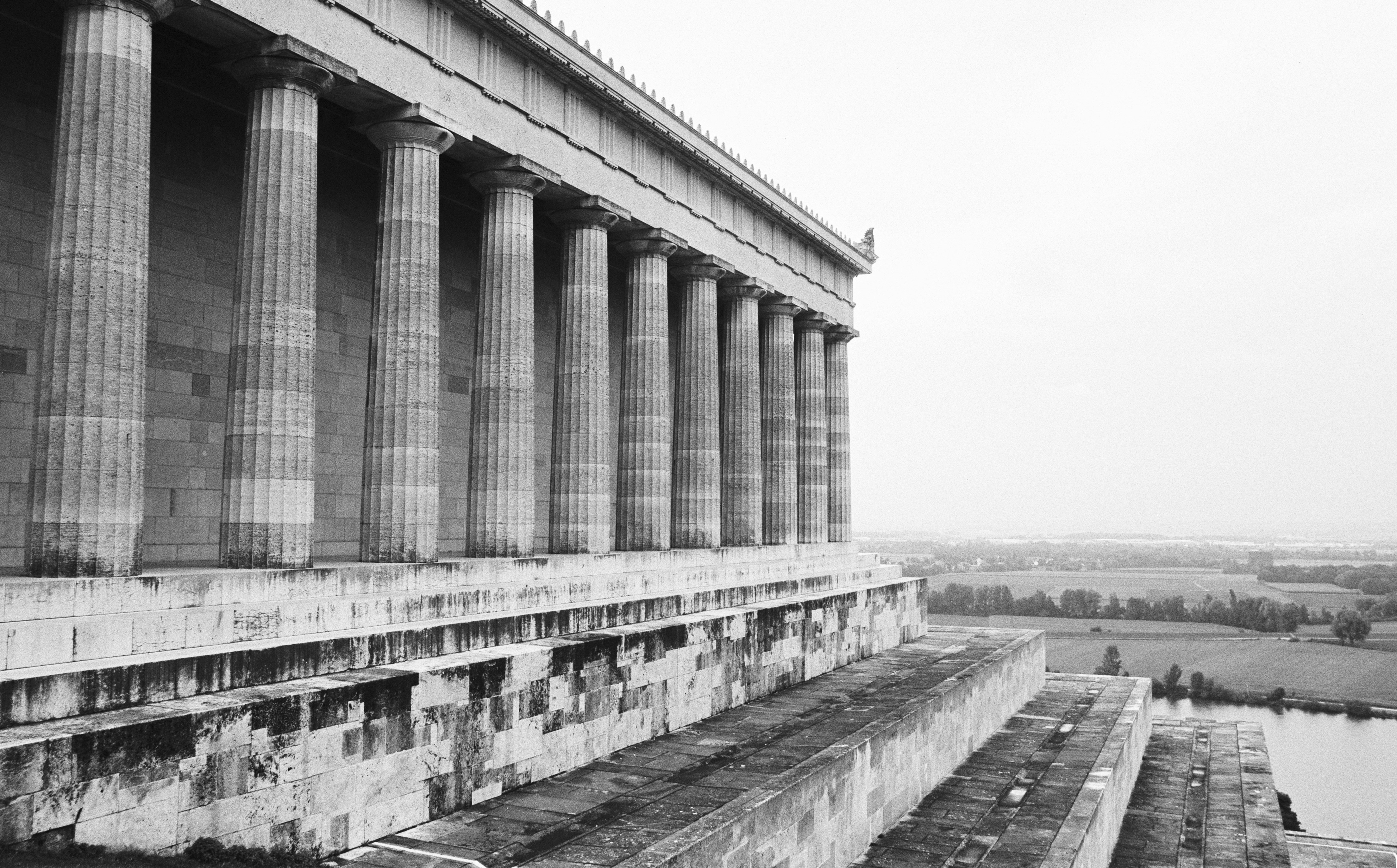 Monochrome architectural photograph of a grand colonnade and stepped terrace, shot from a low angle to emphasize its lines. Weathered textures and the horizon recede into the distance.