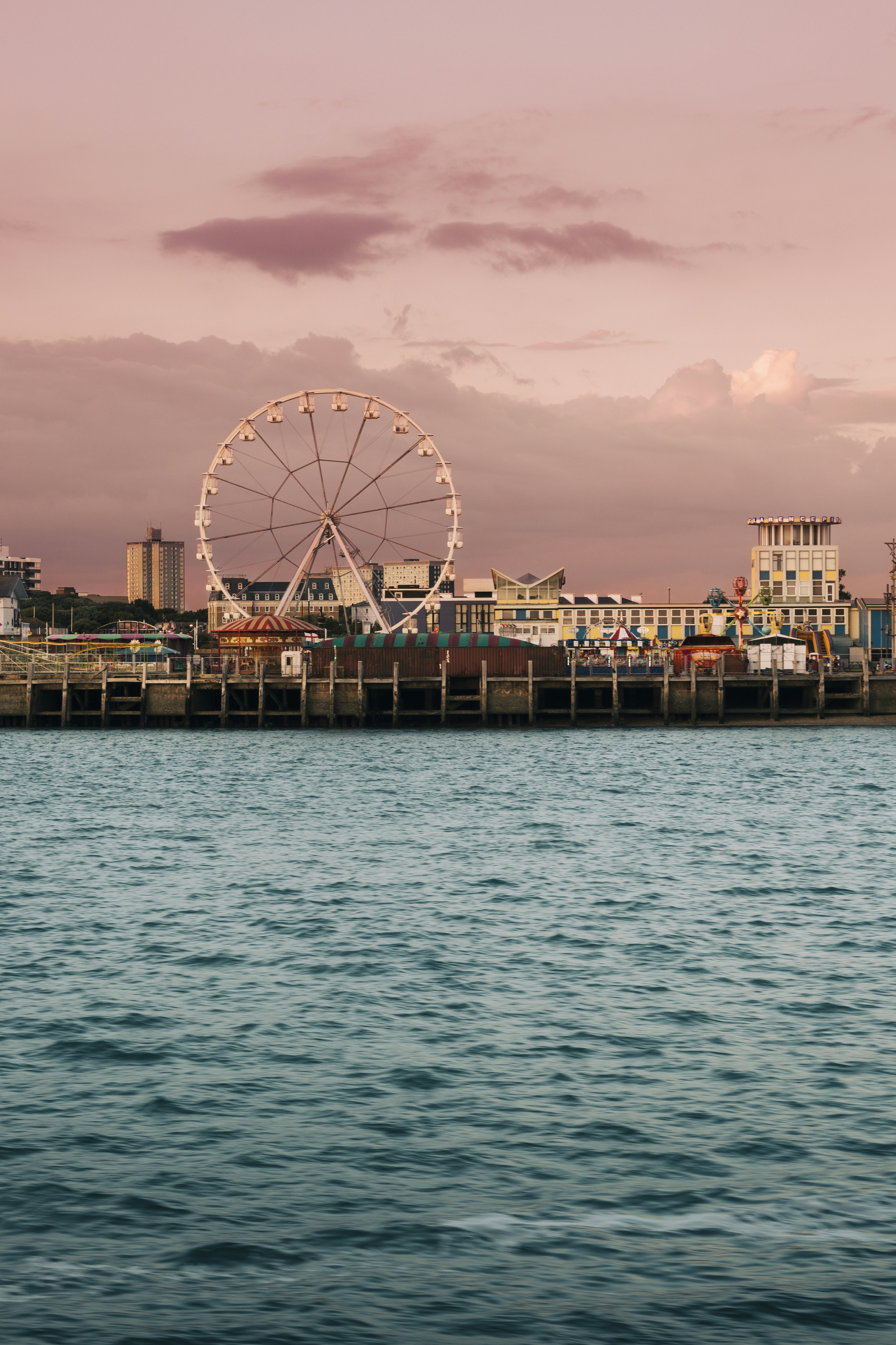 A vibrant Ferris wheel stands tall against a pastel sky, reflecting the lively atmosphere of the waterfront. The scene captures the essence of leisure and enjoyment by the water.