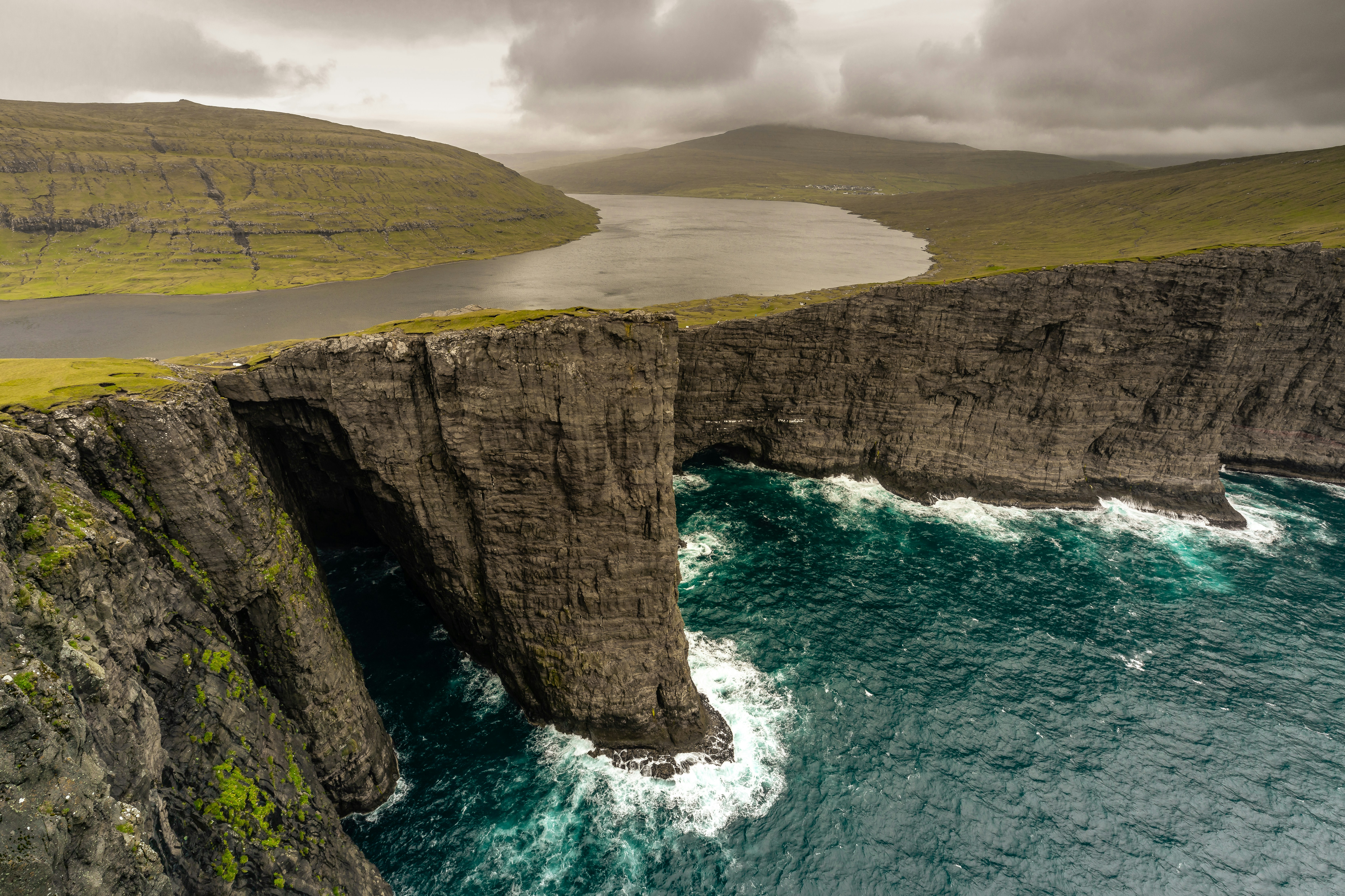 Lake Sørvágsvatn appears to float above the sea cliffs, with turbulent ocean waves below and misty skies above.