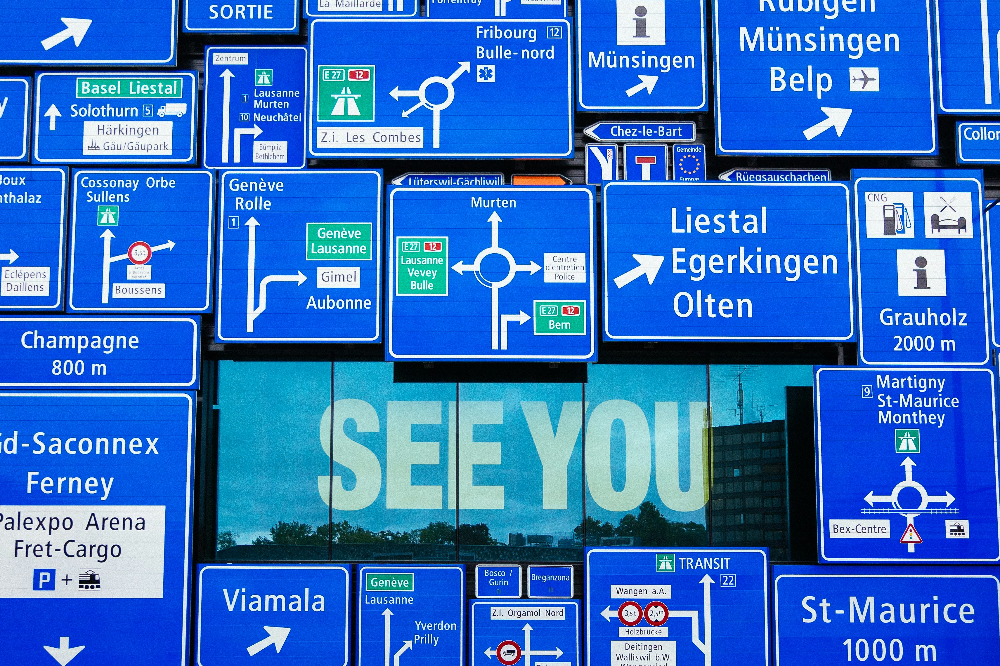 Array of blue road signs with various directions surrounding a glass panel displaying 'SEE YOU'.