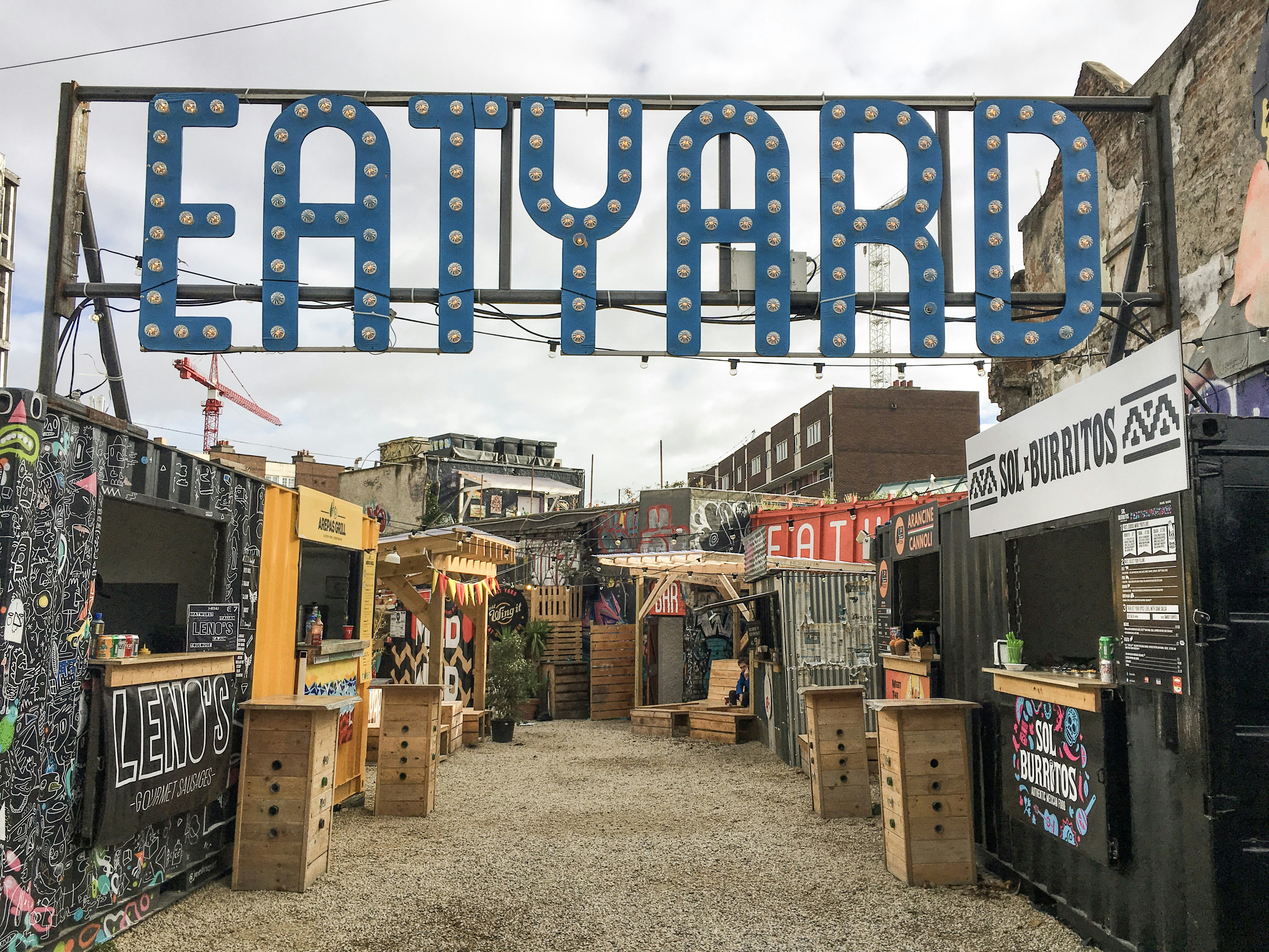 Open-air food market with vibrant stalls under a large marquee sign on a cloudy day.