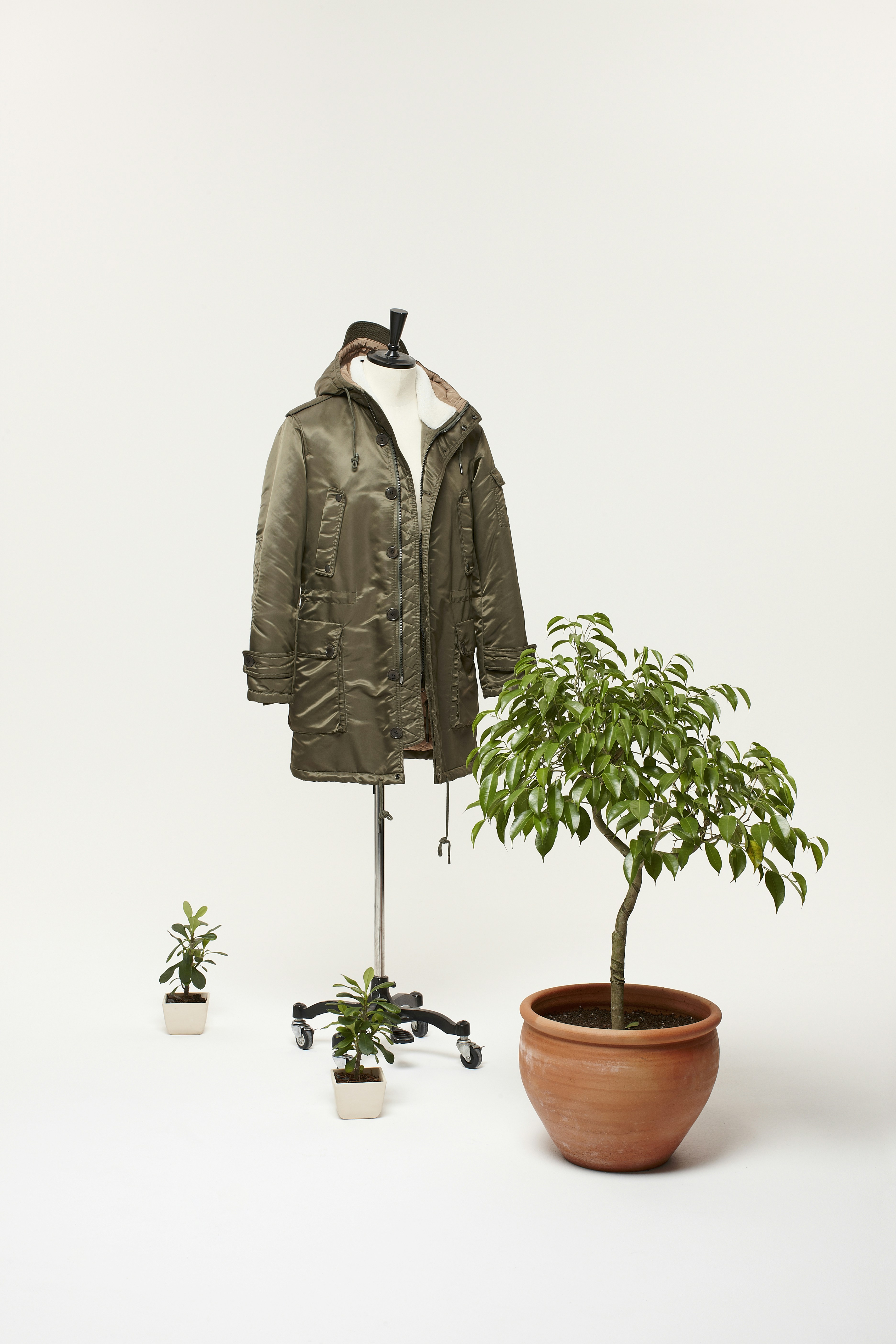 Olive green parka displayed on a mannequin beside potted plants in a minimalist setting.