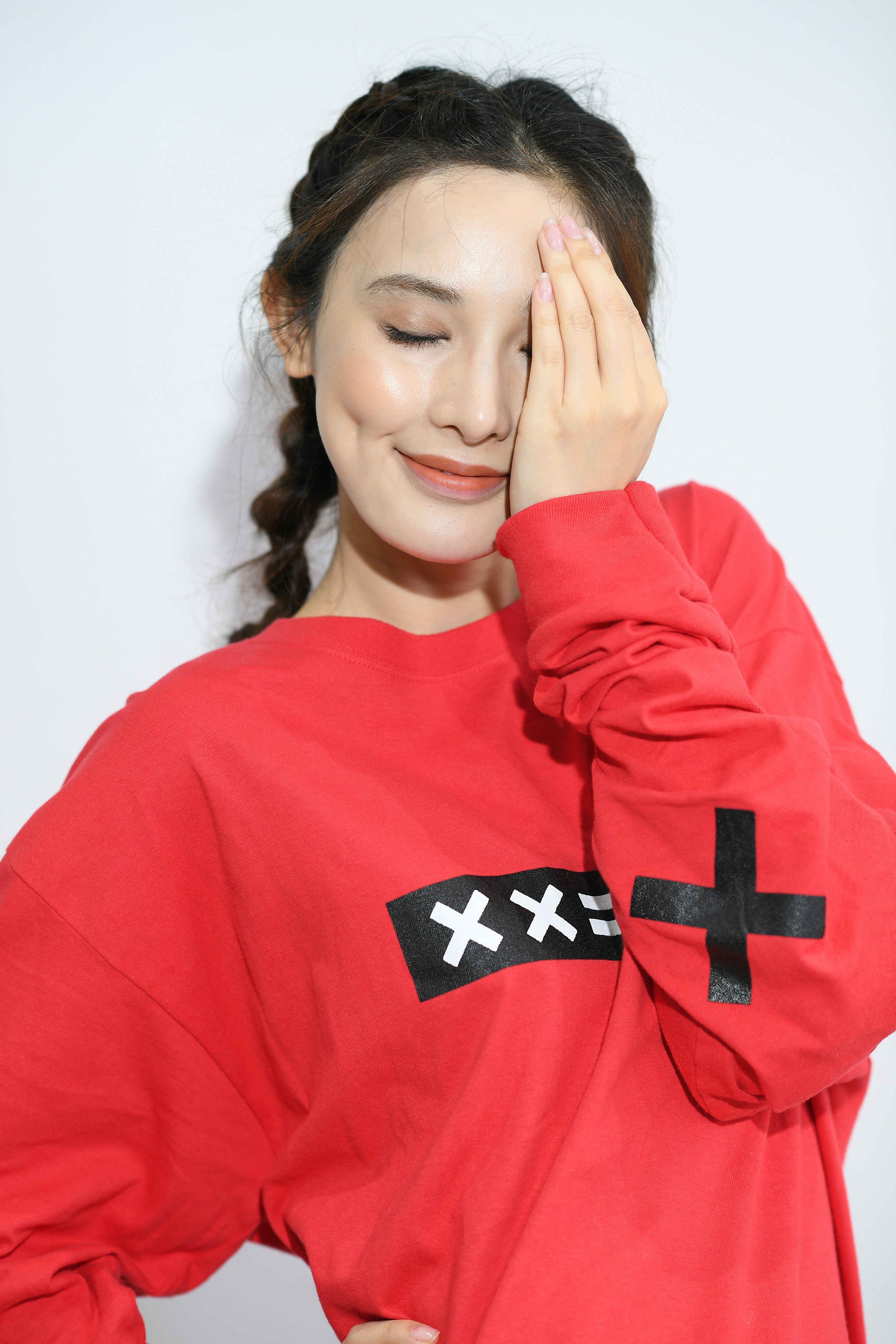 Young woman in a red sweatshirt covering one eye with her hand, smiling softly against a plain white background.