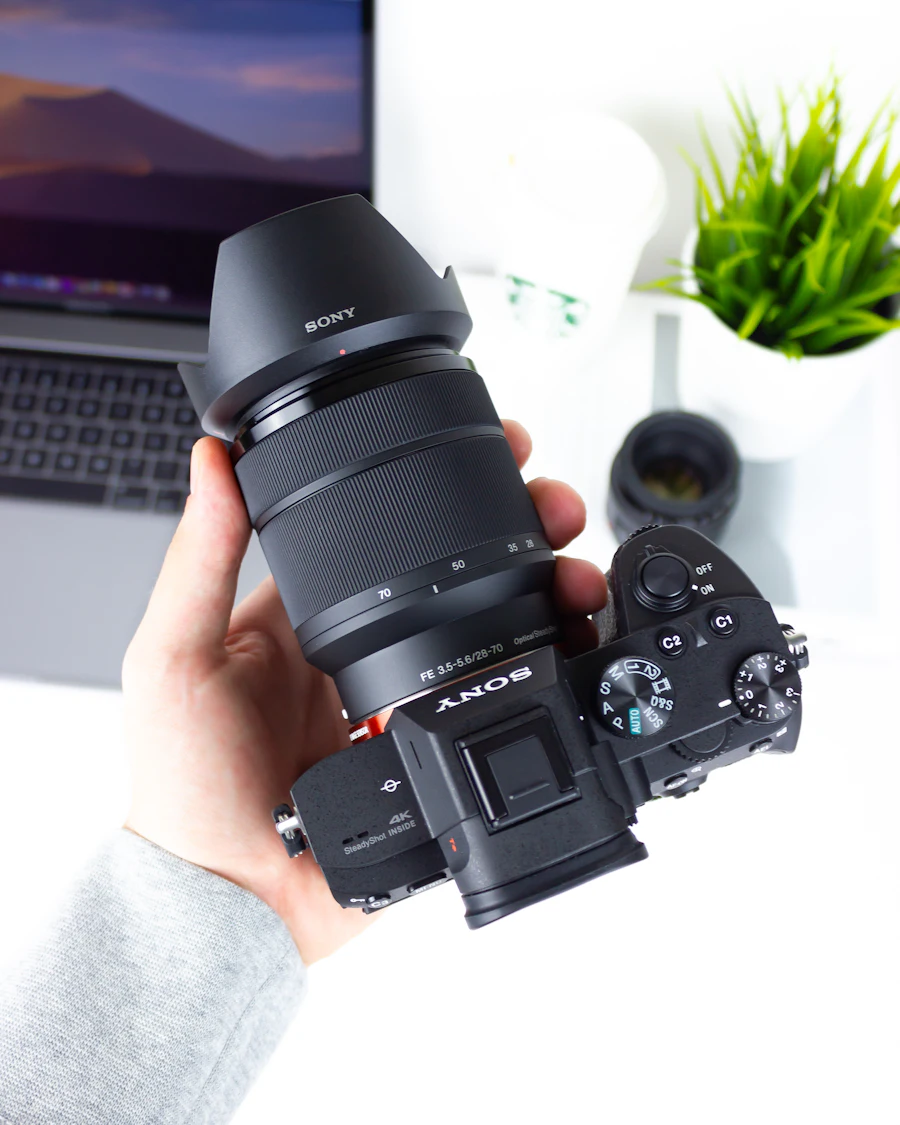 A hand holds a Sony mirrorless camera fitted with a large zoom lens on a bright desk beside a laptop and a potted plant.