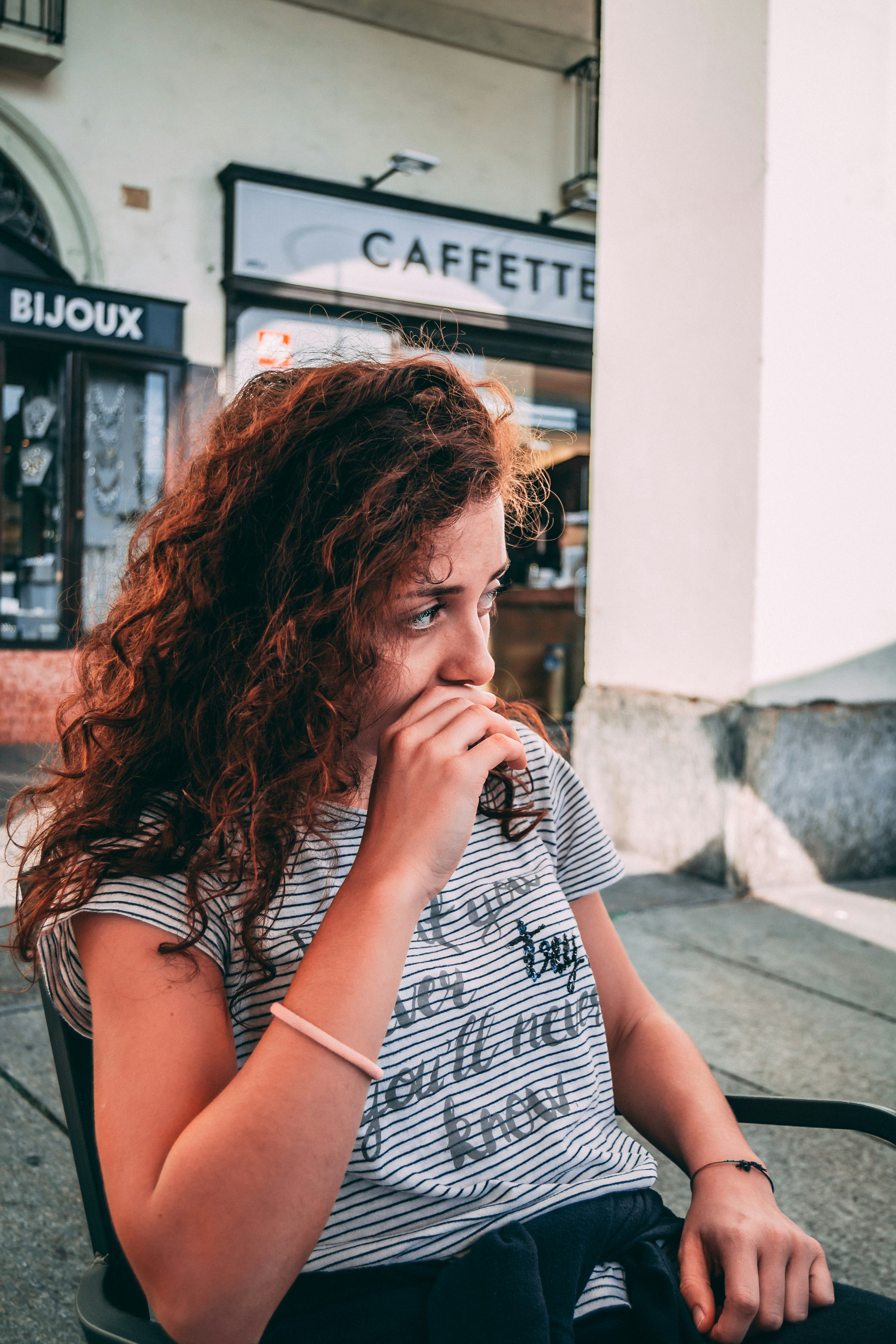 Person with curly hair seated outdoors, gazing thoughtfully towards the street.