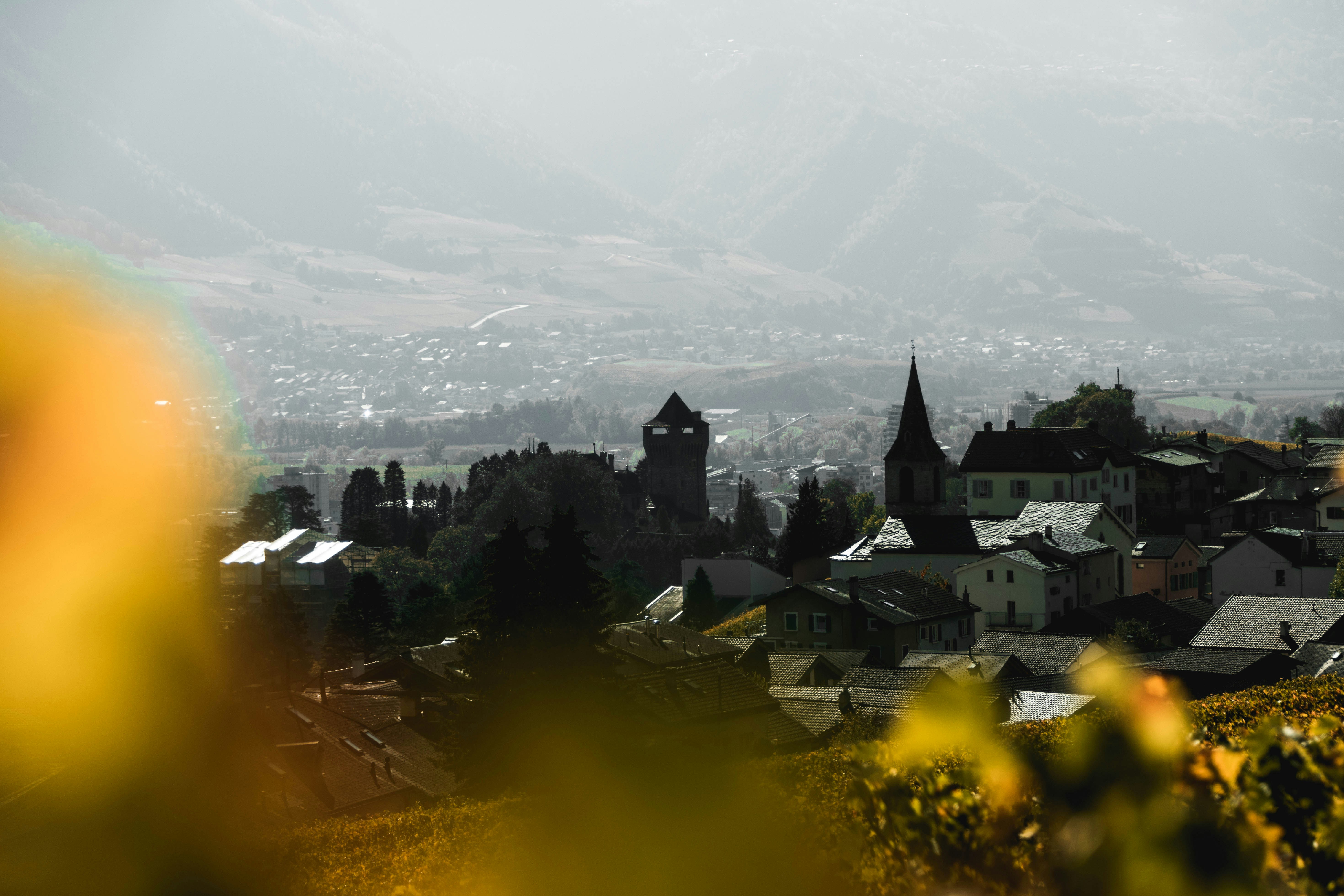 Historic village nestled in a valley, framed by distant mountains and soft foreground foliage. A blend of architecture highlights the serene landscape.