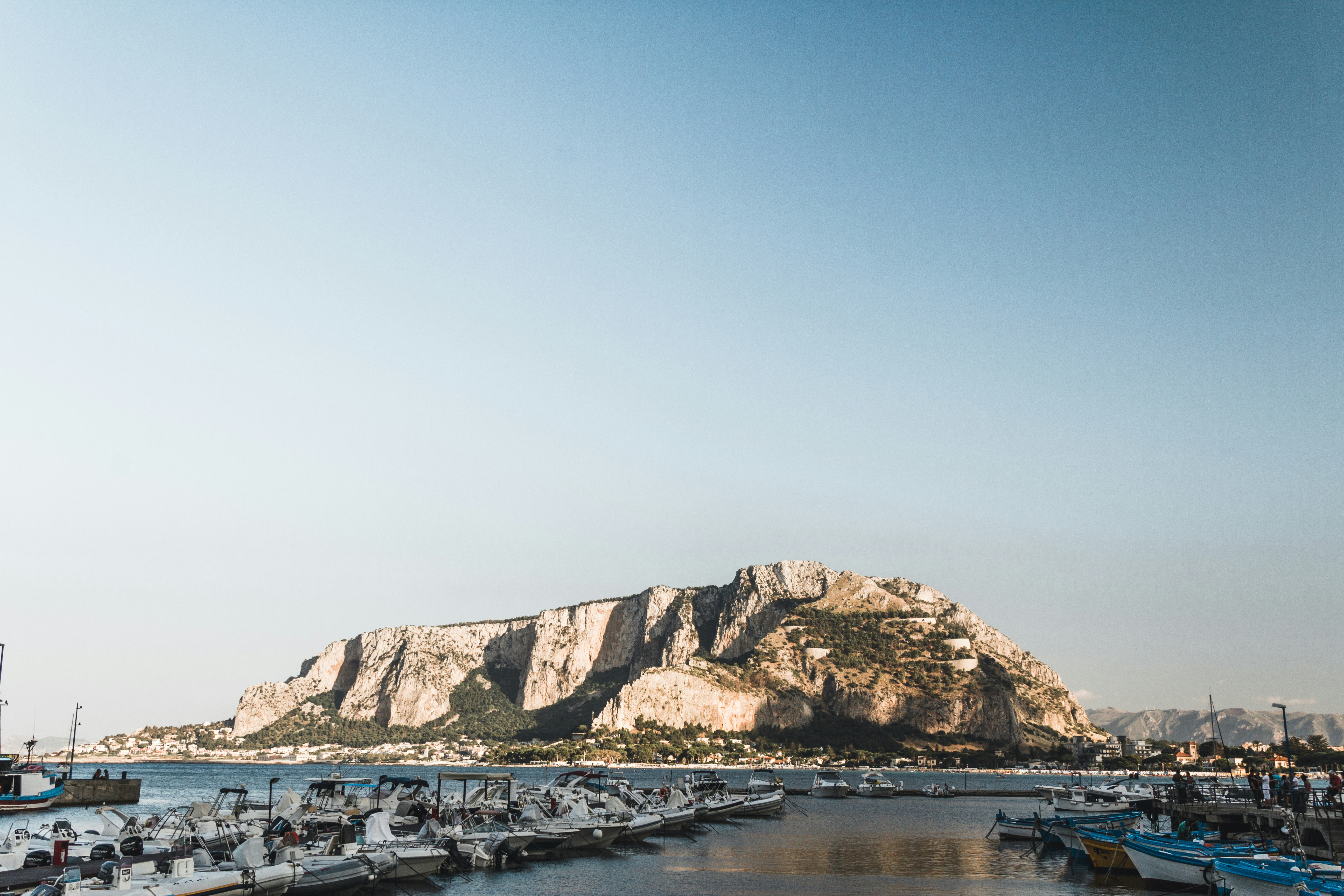 Rocky hill illuminated by sunlight, framed by a serene marina filled with boats under a clear blue sky.