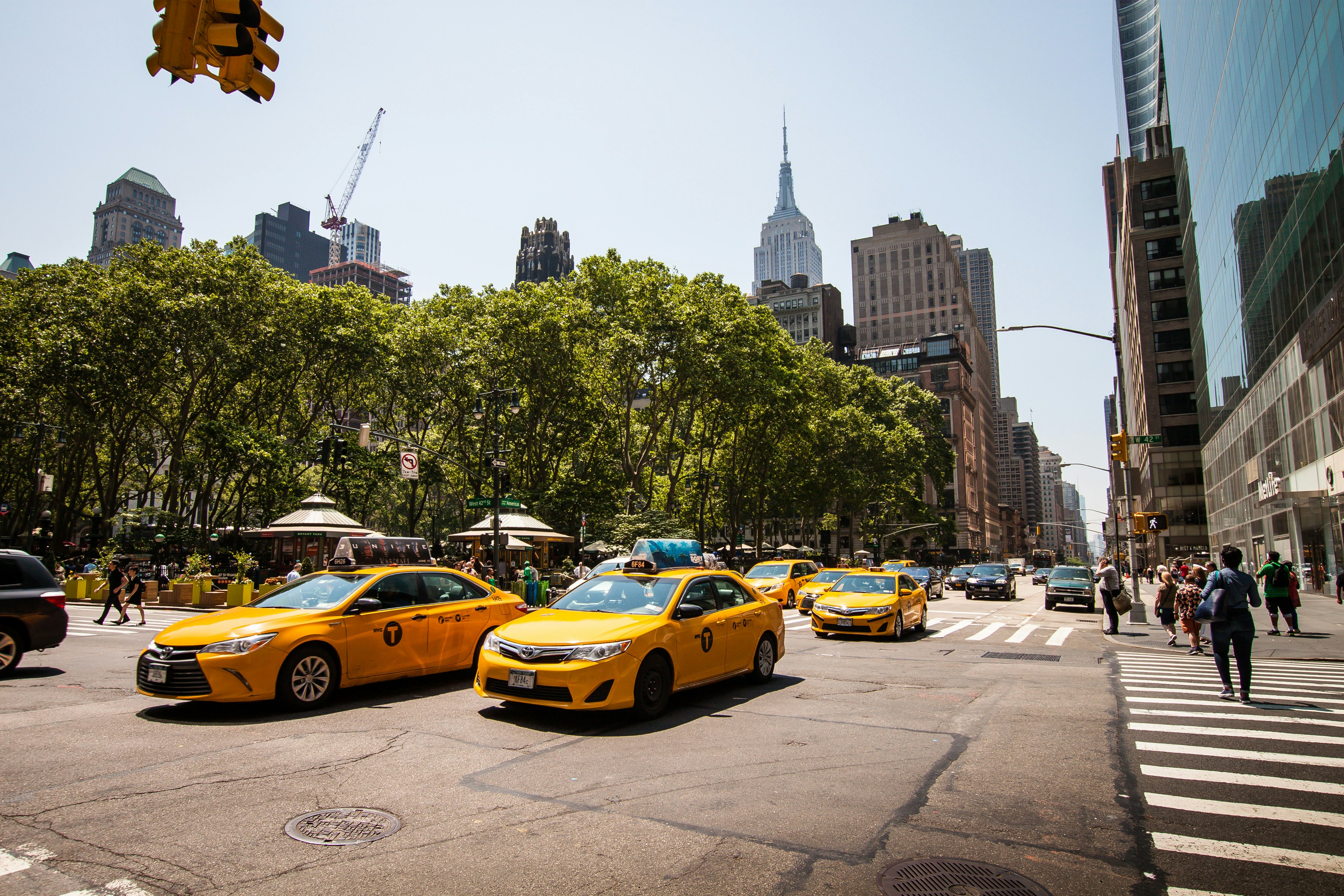 Yellow taxis navigate a bustling city intersection with skyscrapers and greenery in the background. The iconic Empire State Building stands tall amidst the urban landscape.
