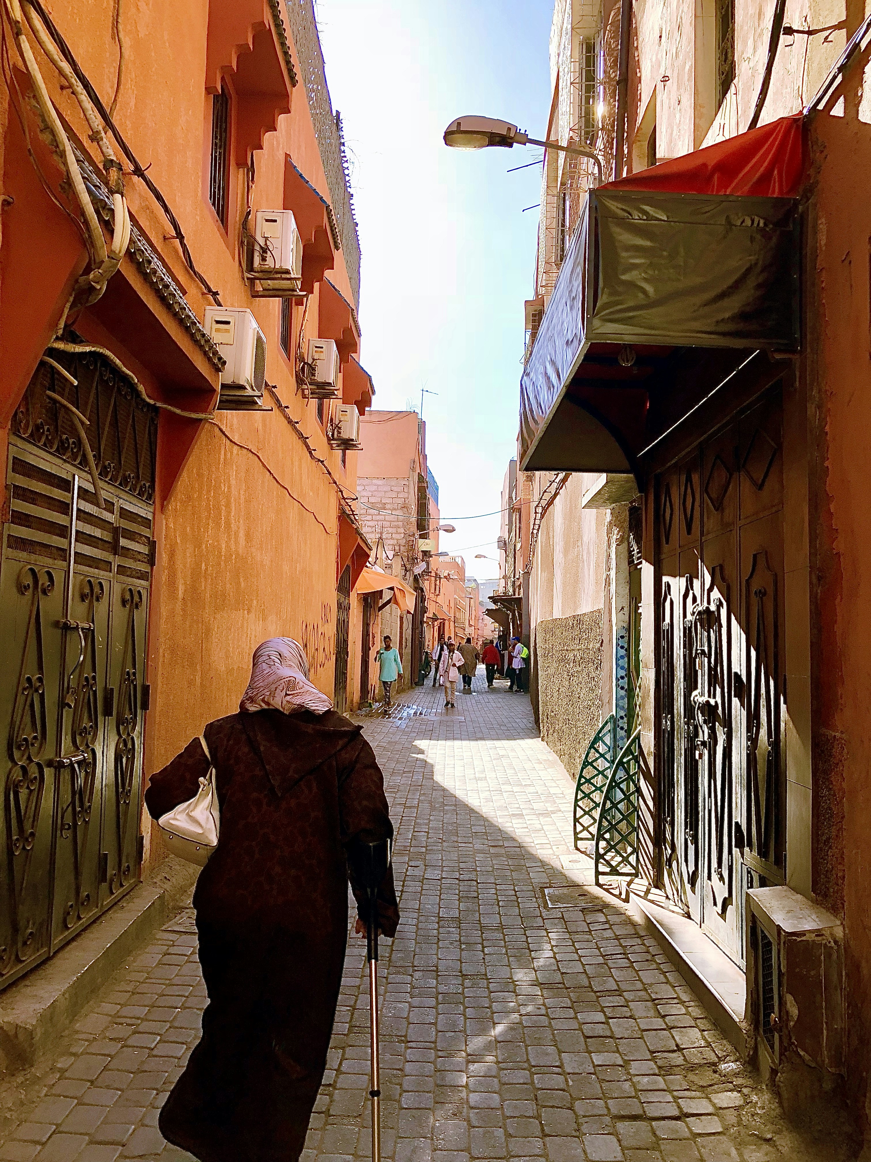 A woman in traditional attire walks down a narrow alleyway, flanked by vibrant orange walls and shadows cast by overhead structures.