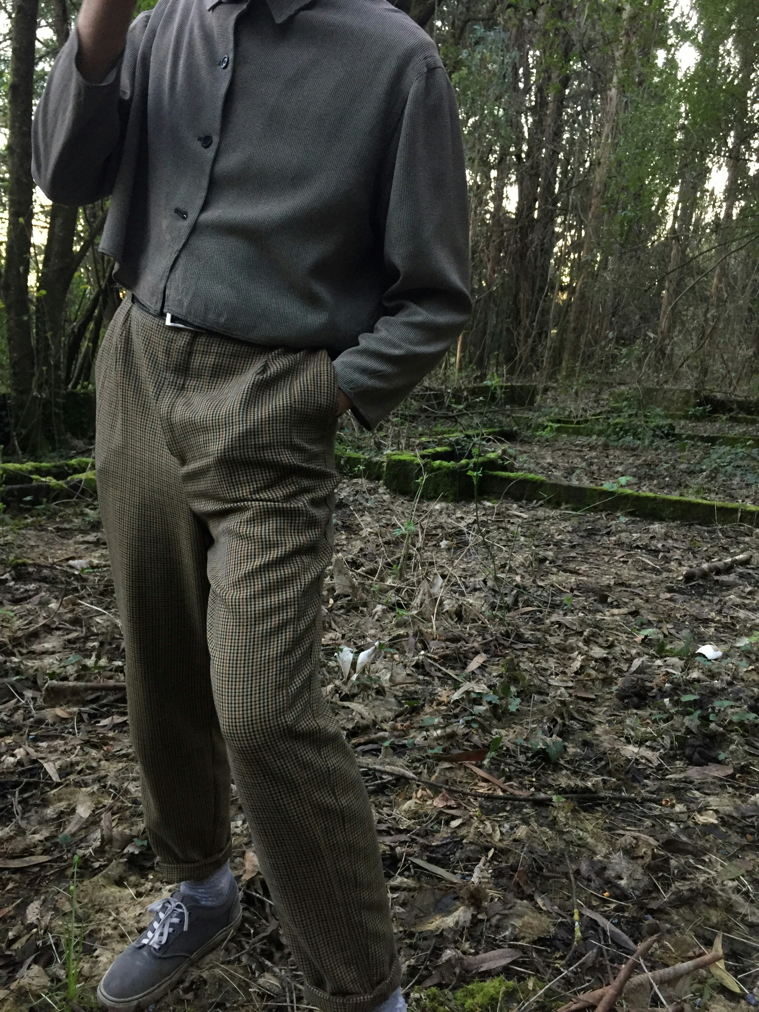 Person in earth-toned attire standing in a forest clearing with soft, diffused light filtering through trees.
