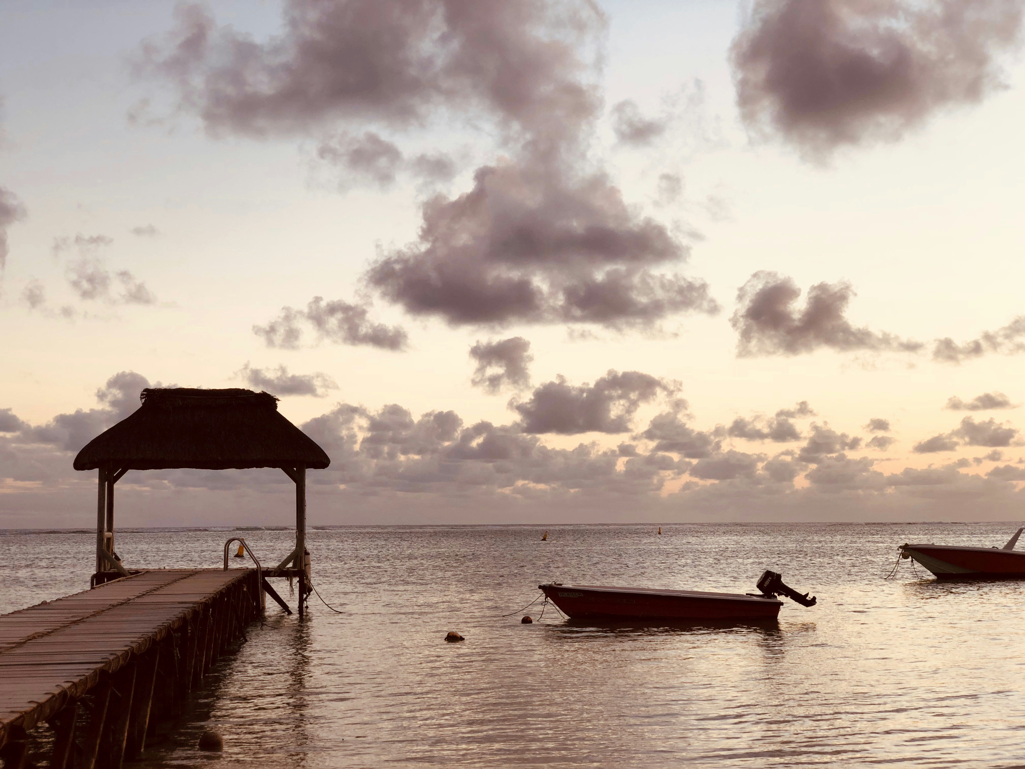 A tranquil scene featuring a wooden dock and boats under a colorful sky at sunset. The calm waters reflect the soft hues of the evening.