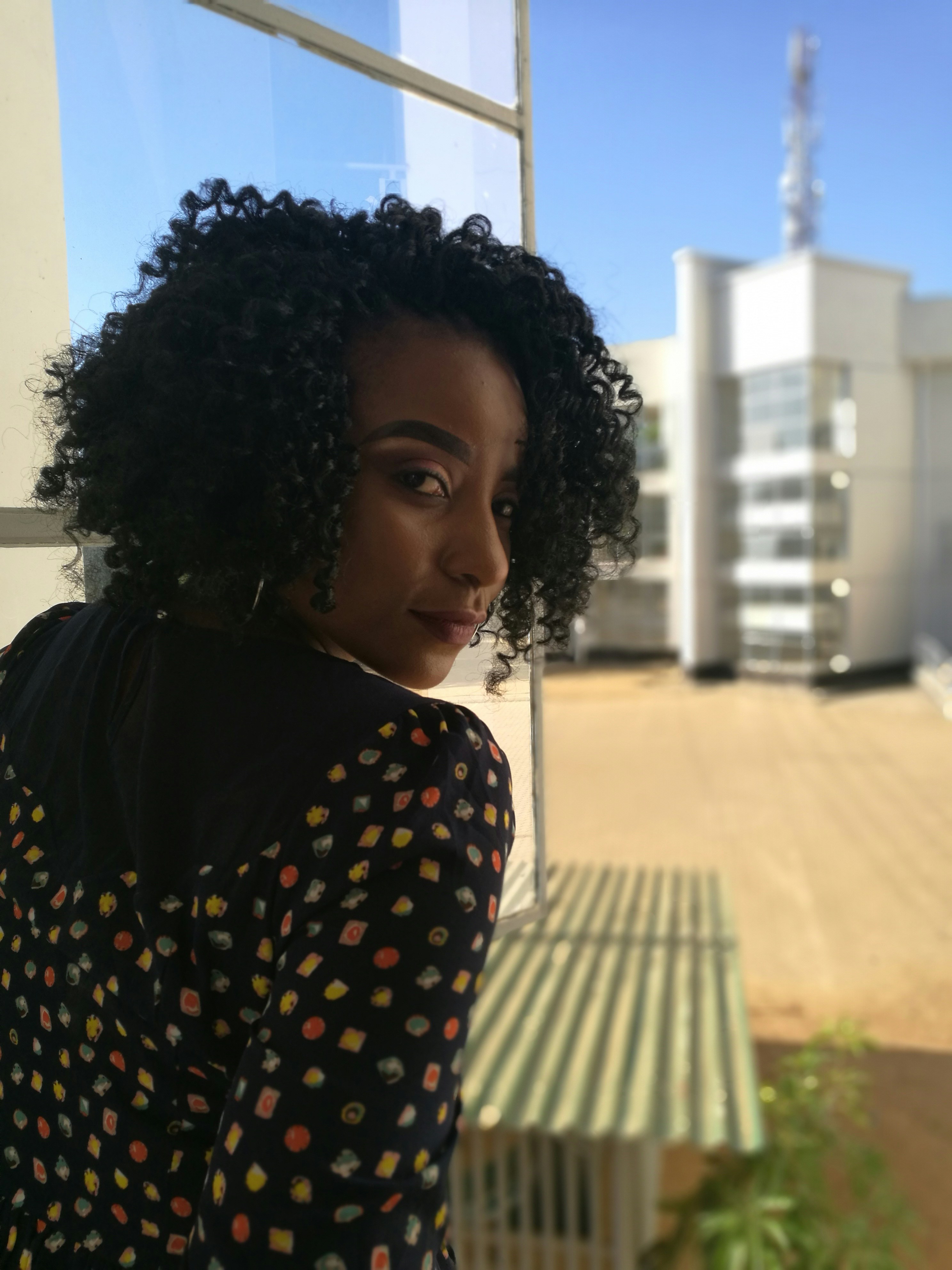 Young woman with curly hair glancing back while standing by a window, showcasing a blend of indoor and outdoor elements. 