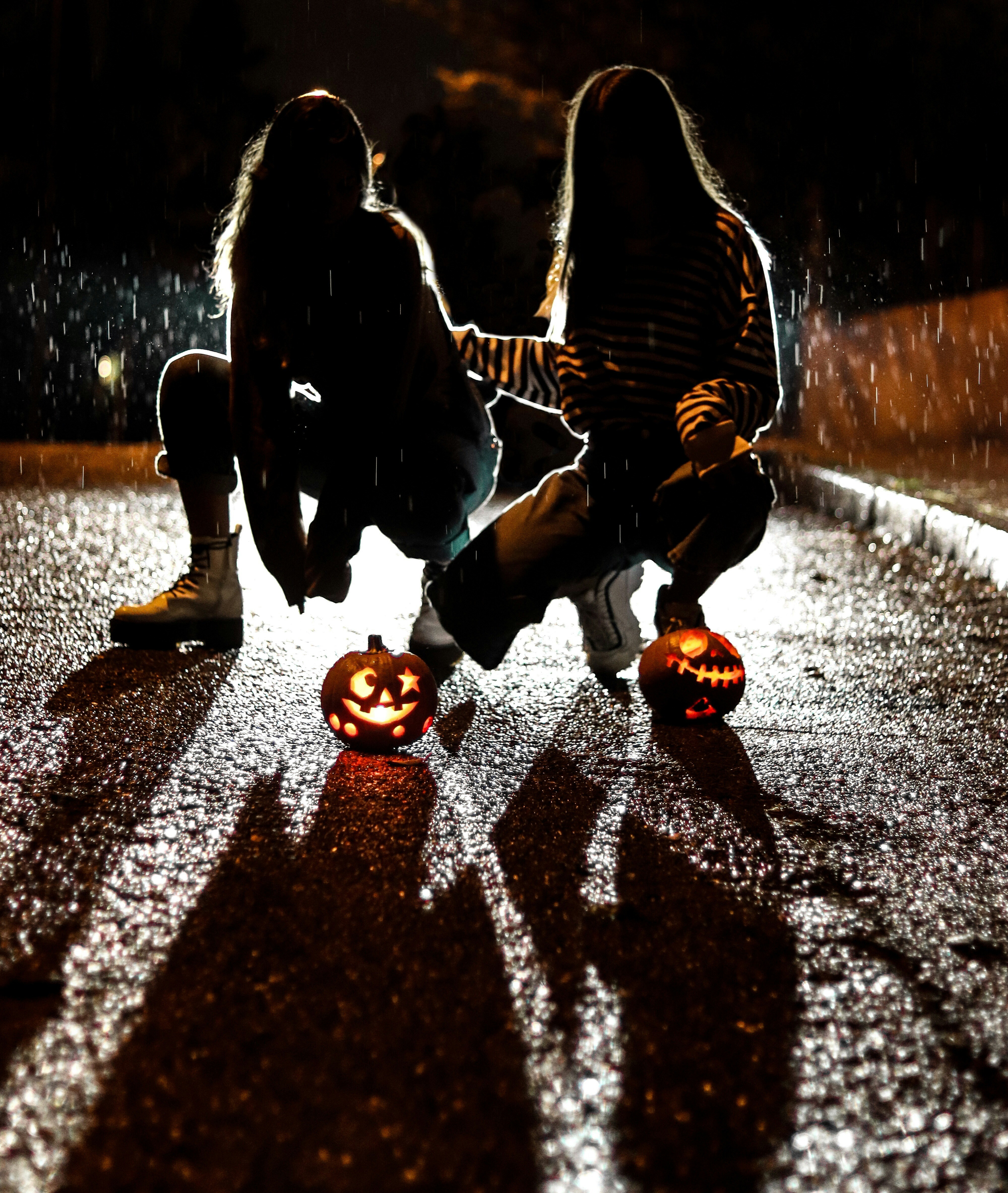 Two figures crouch on a rain-slicked street, illuminated by the eerie glow of carved pumpkins. The scene captures a moment of playful spookiness amidst the darkness.